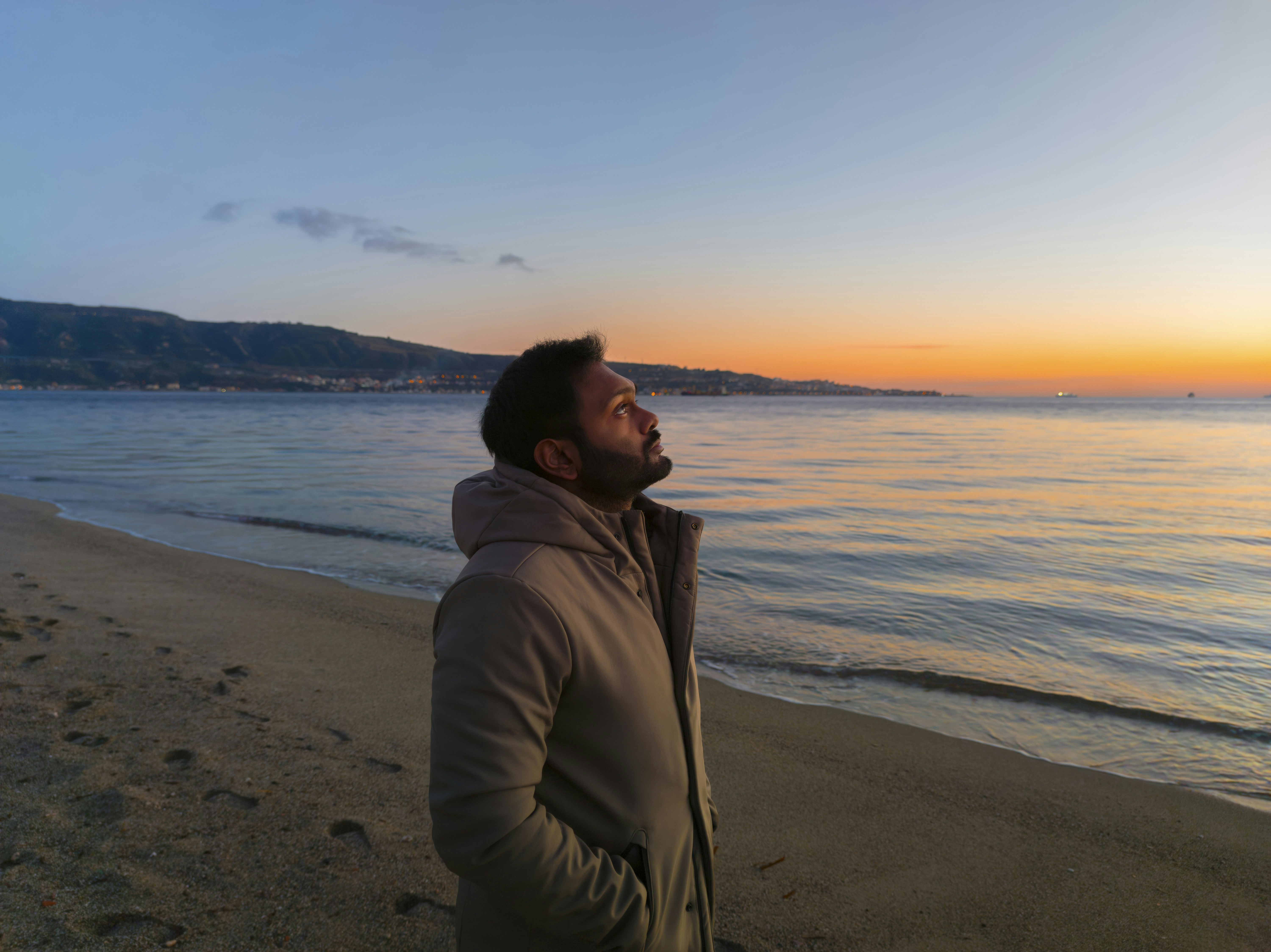 A man in a tan coat stands on a sandy beach at sunset, gazing toward the horizon as waves roll in.
