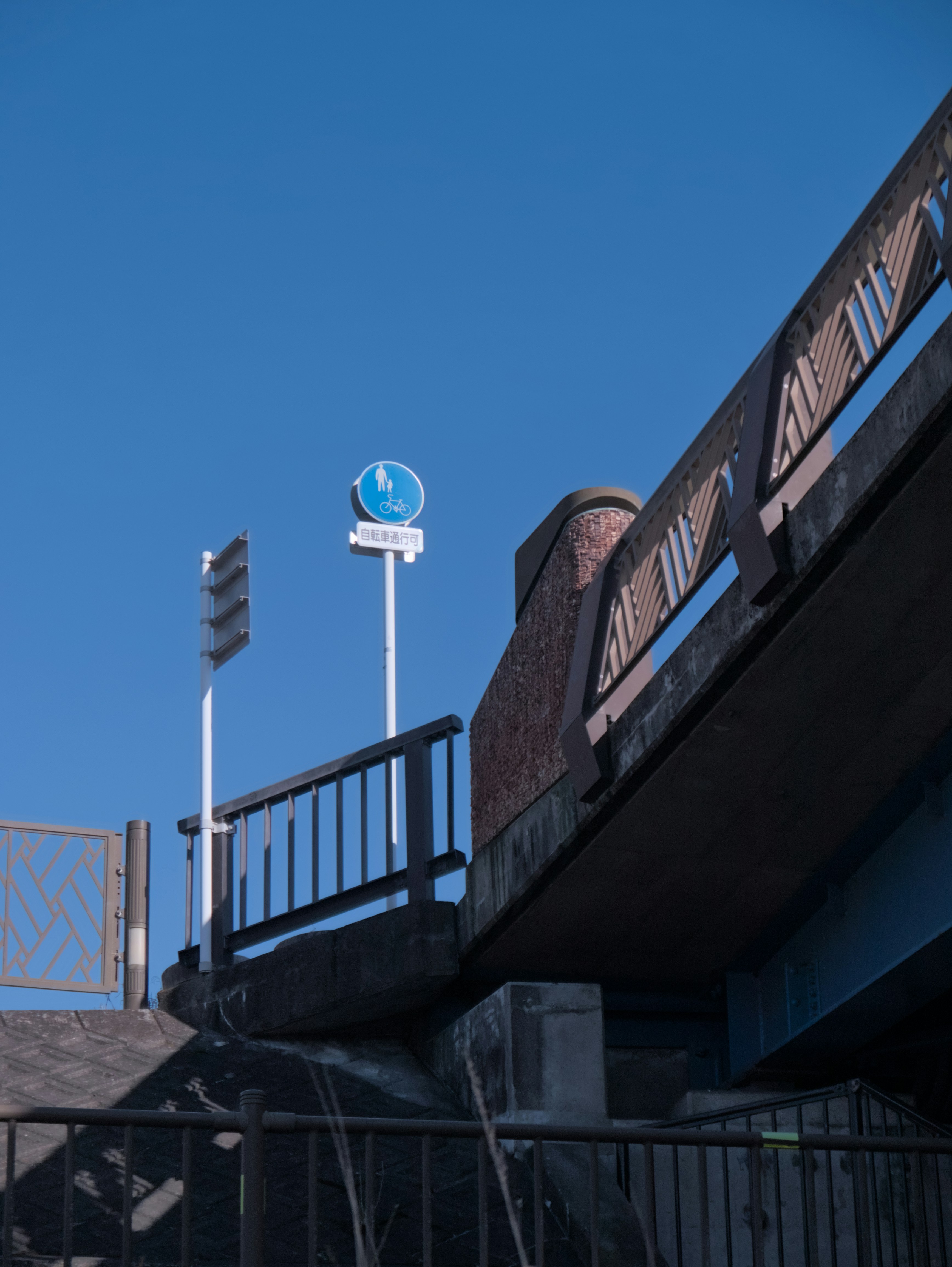 Blue Bicycle Route Sign Above an Angular Urban Bridge Against a Crisp Blue Sky