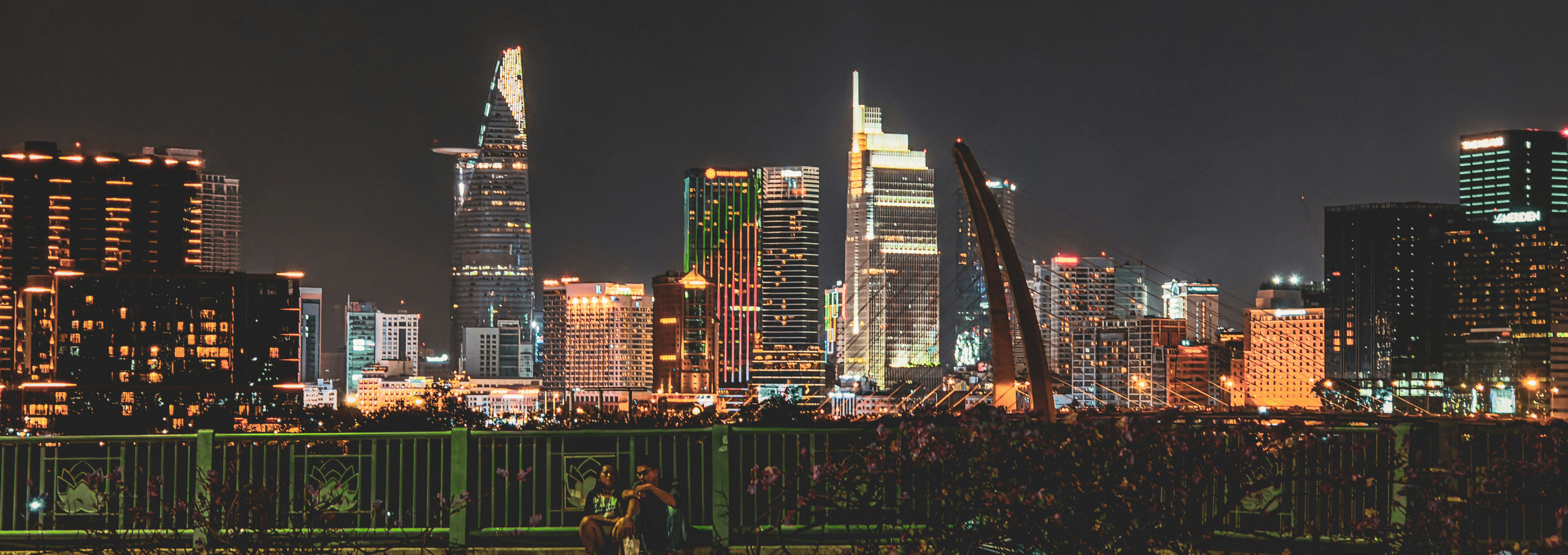 Vibrant city skyline illuminated at night, showcasing modern architecture and dynamic lighting. The foreground features a green railing with silhouettes of people enjoying the view.