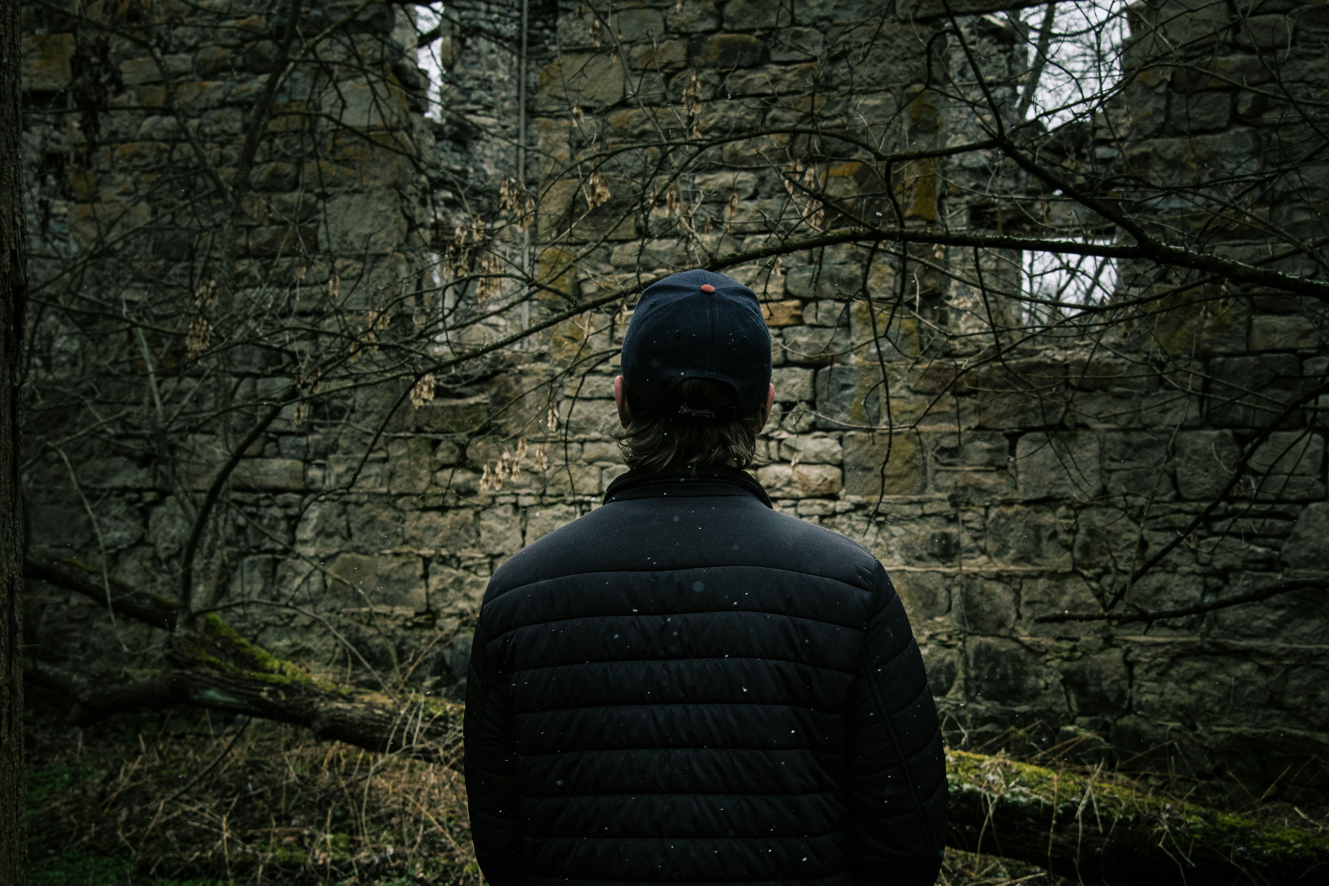 Person in a dark jacket and cap standing before weathered stone ruins surrounded by overgrown vegetation.