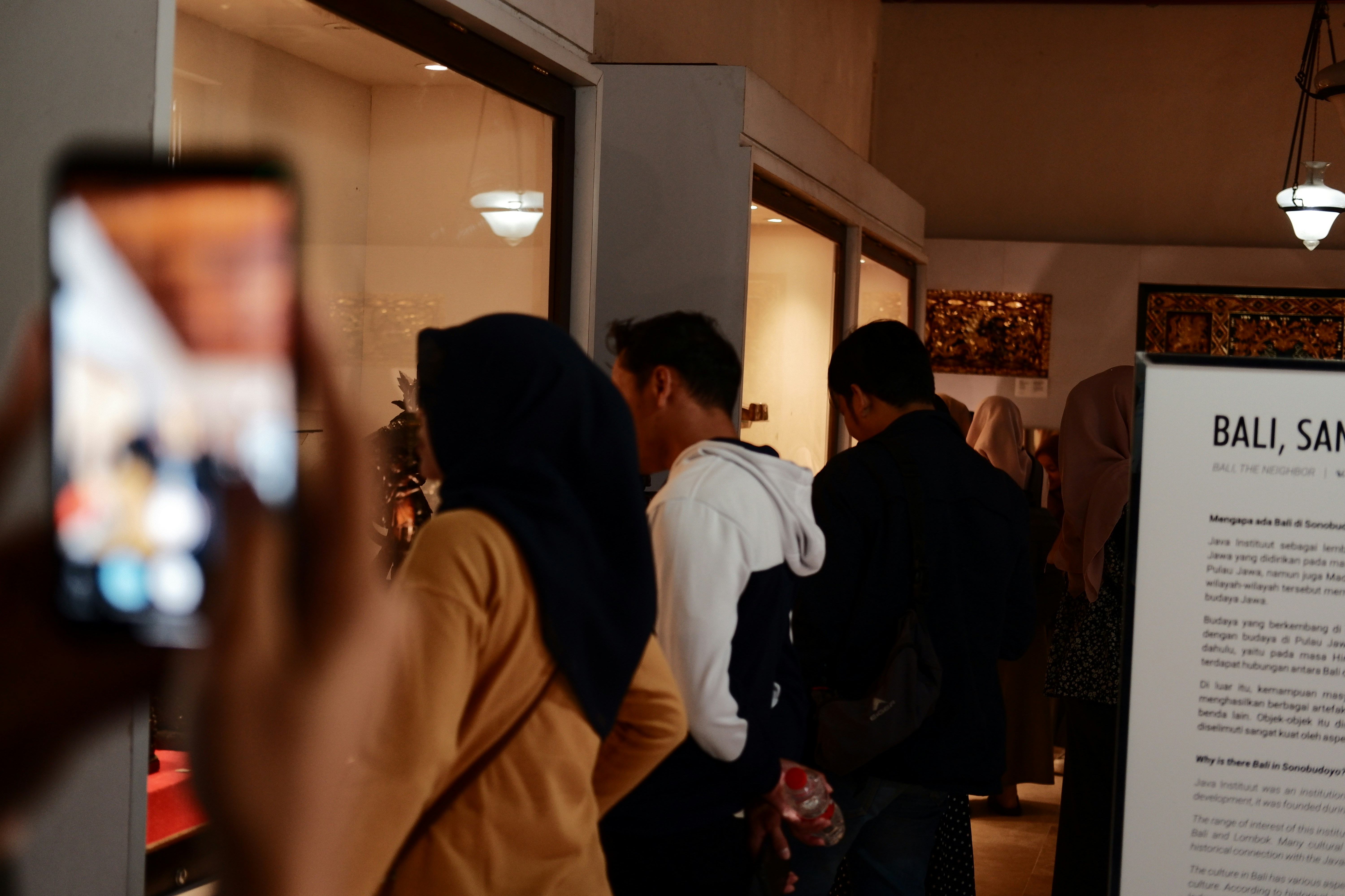 Visitors engaged in exploring Balinese artifacts in a museum setting, with a smartphone capturing the moment. 