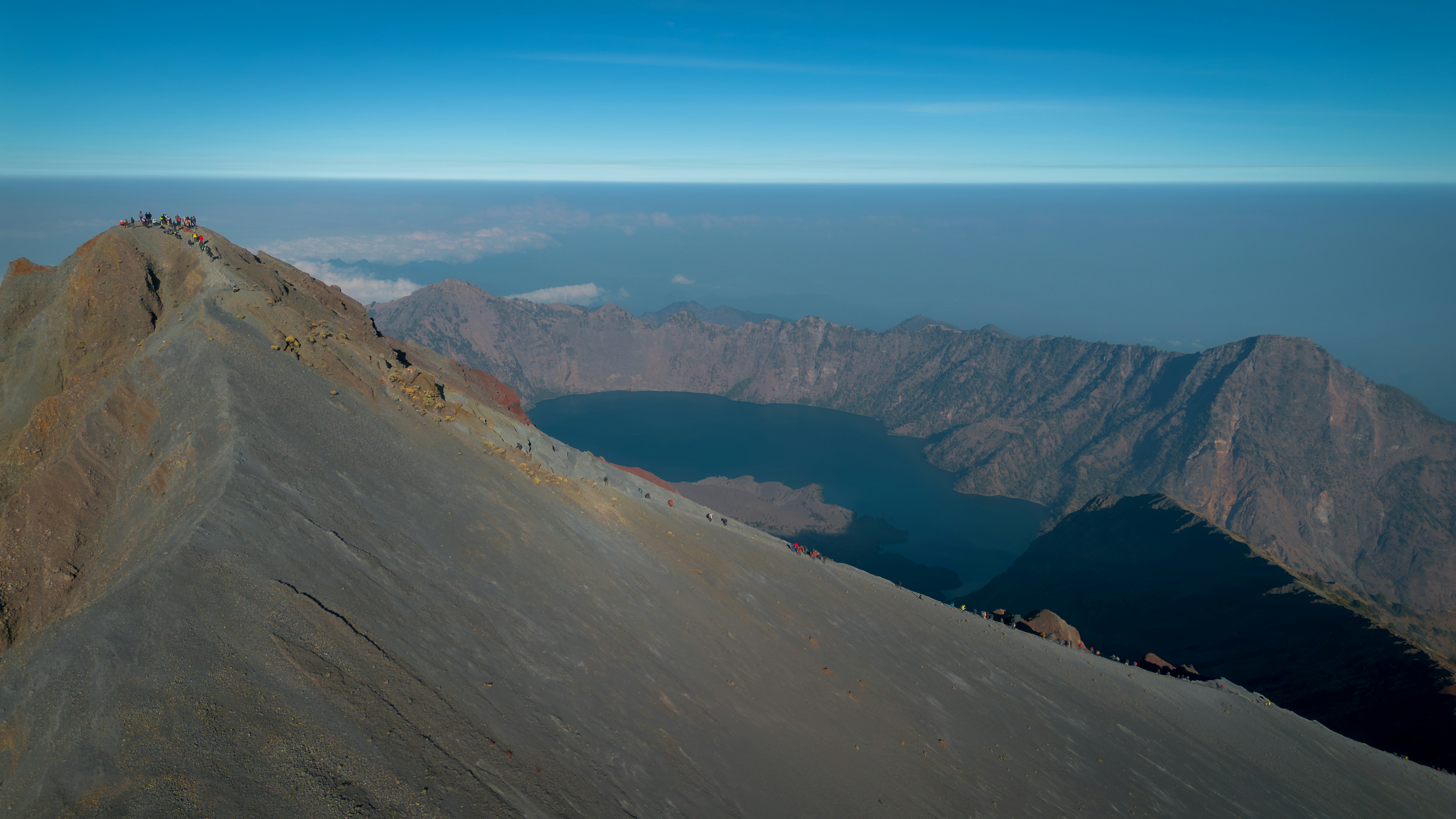 Hikers traverse the rugged slopes of Mount Rinjani with the crater lake Segara Anak in the distance.
