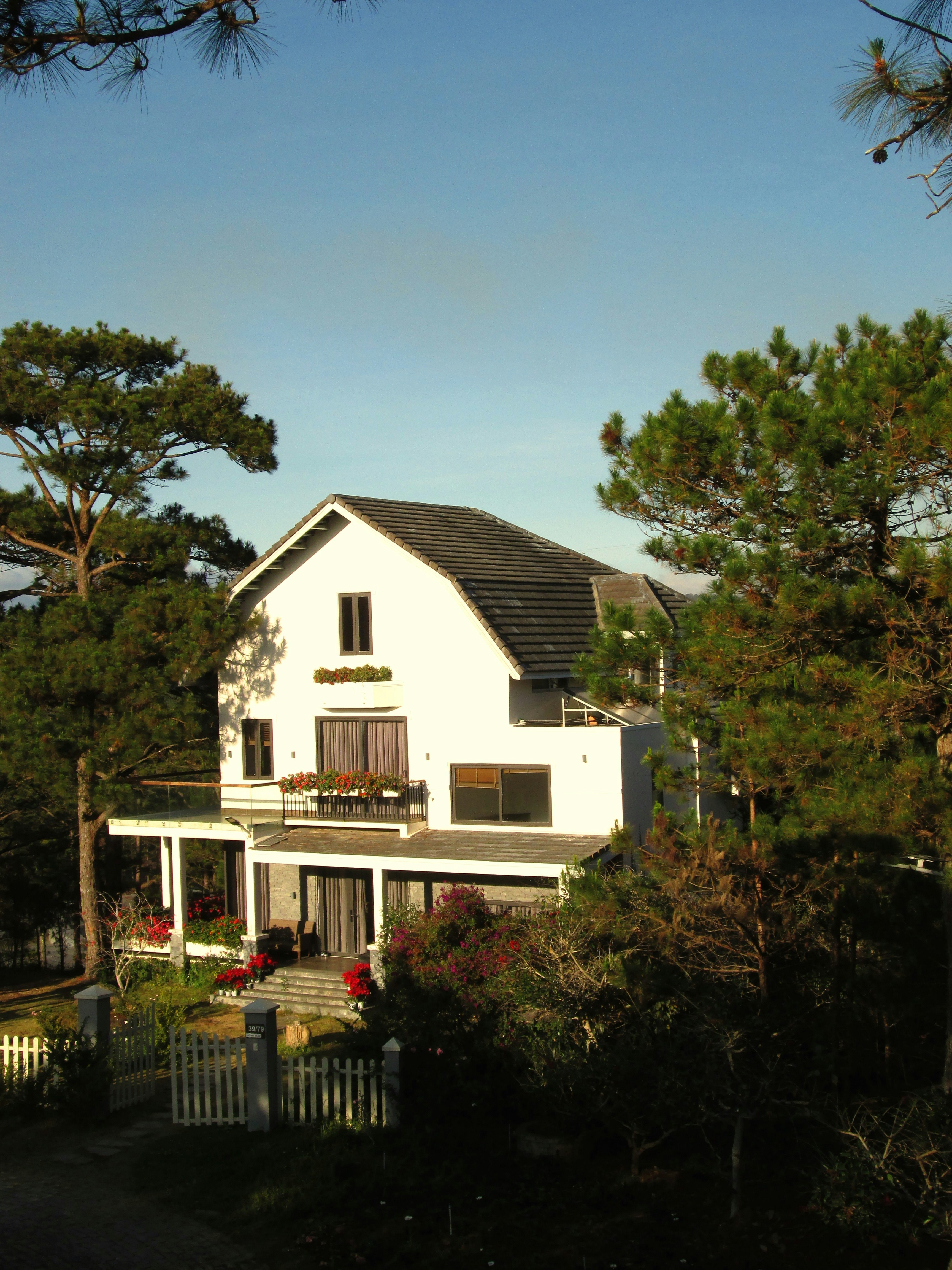 Two-story white house with a dark shingled roof sits among tall pines, featuring a front terrace and blooming planters. A white picket-style fence encloses a well-kept garden under a bright blue sky.