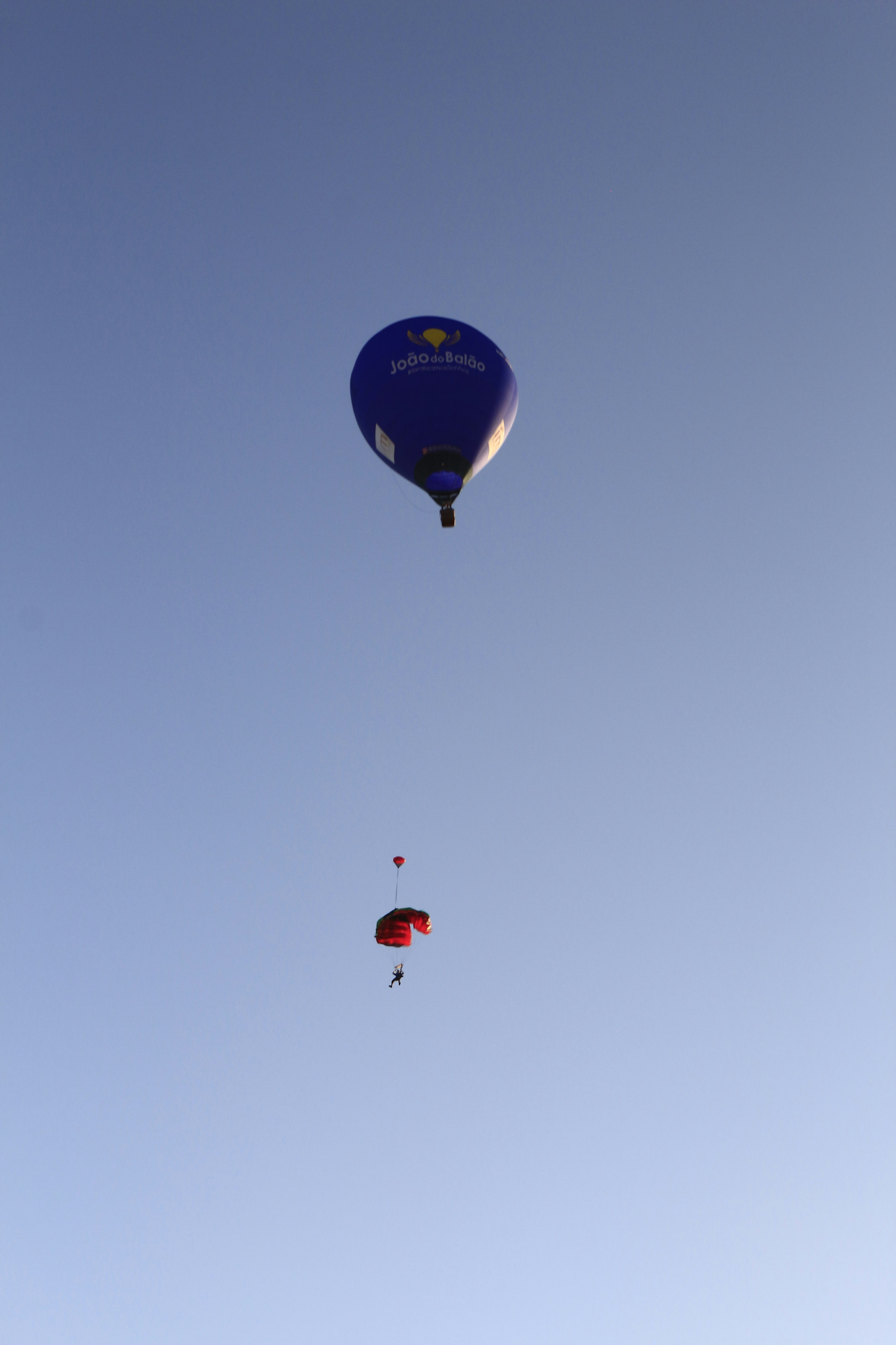 A vibrant hot air balloon floats serenely above, while a parachutist descends gracefully below, creating a striking juxtaposition in the clear blue sky.