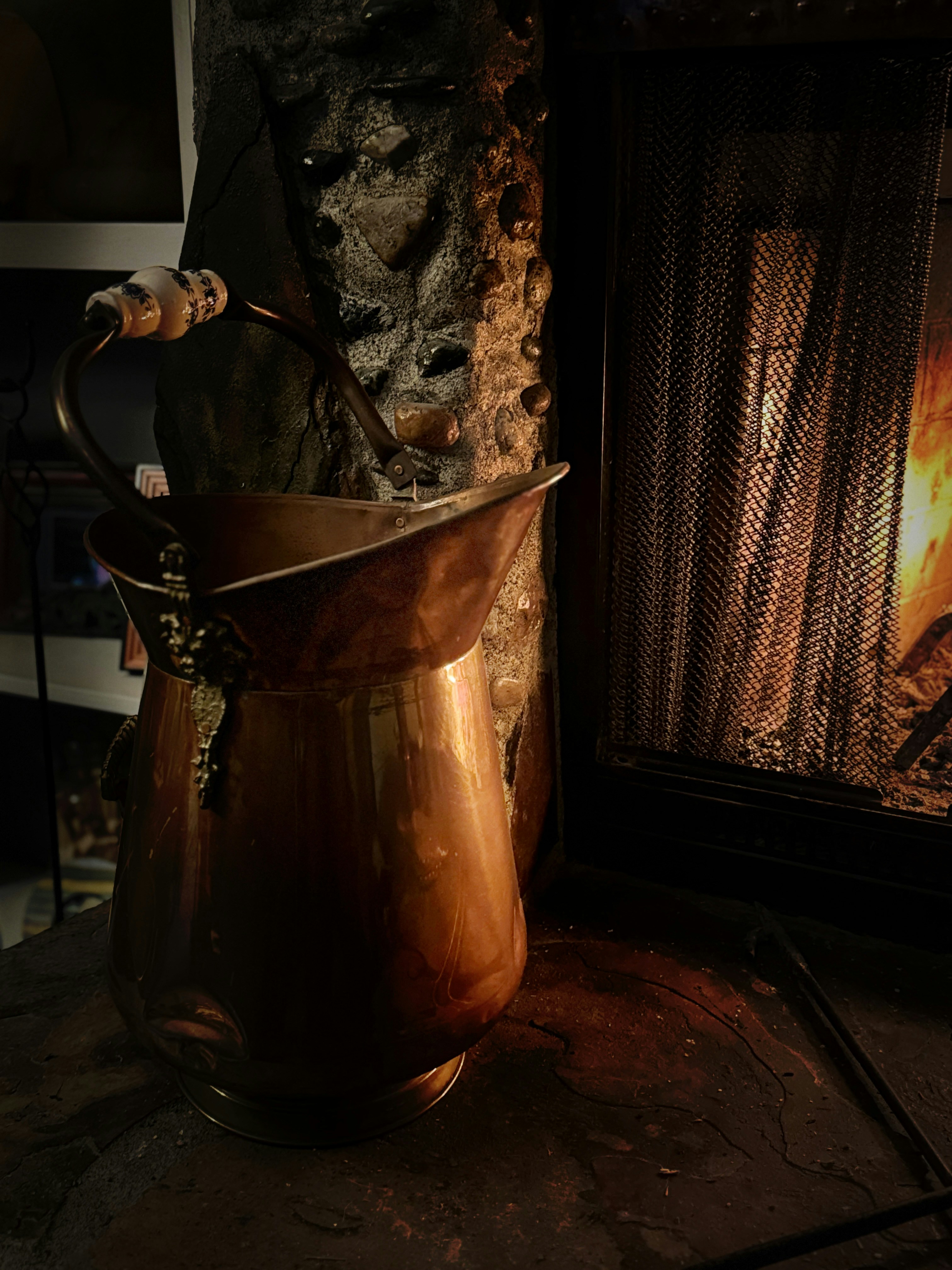 Antique copper pitcher resting beside a glowing fireplace, showcasing the interplay of light and shadow in a cozy setting.