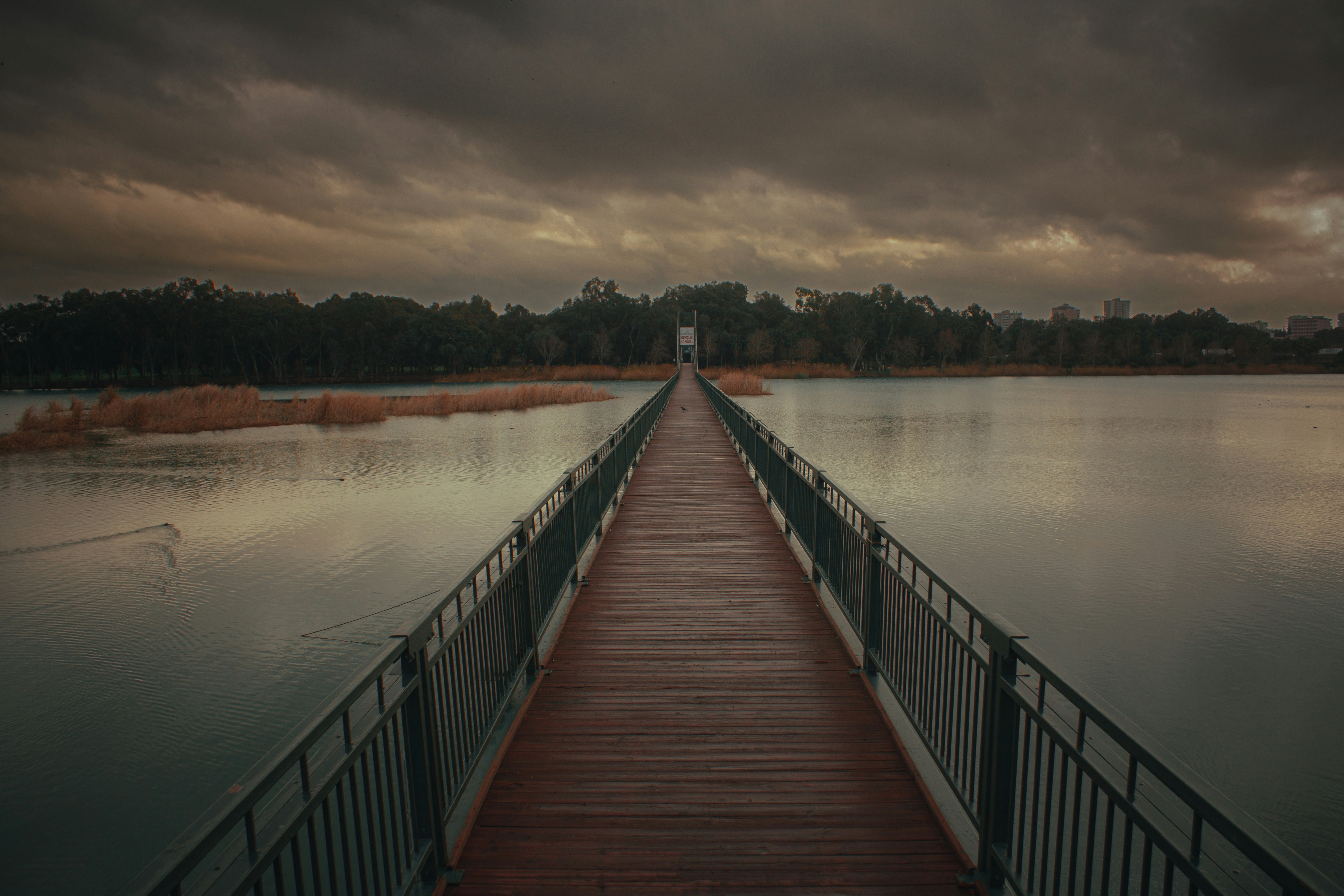 Long wooden bridge stretches across tranquil water under a moody, overcast sky.