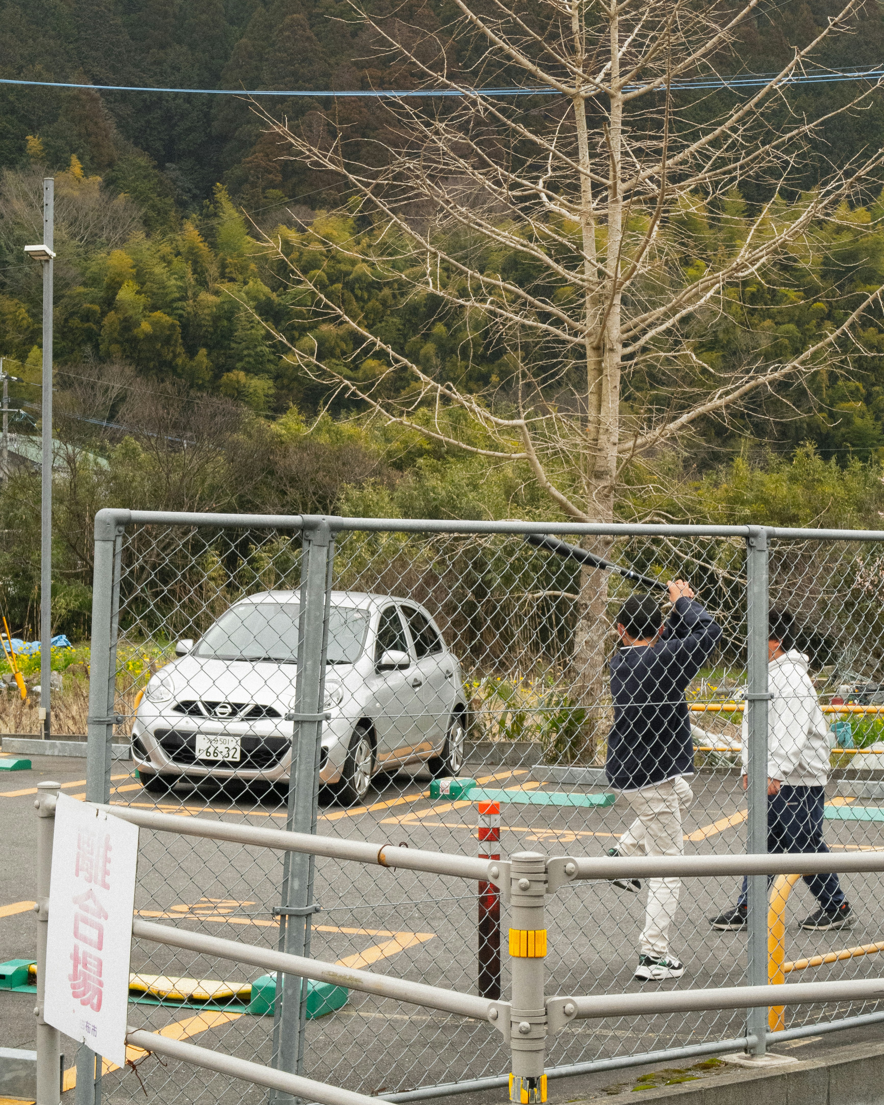 Chain-Link Pause: Two Figures Beside a White Hatchback in a Quiet Lot