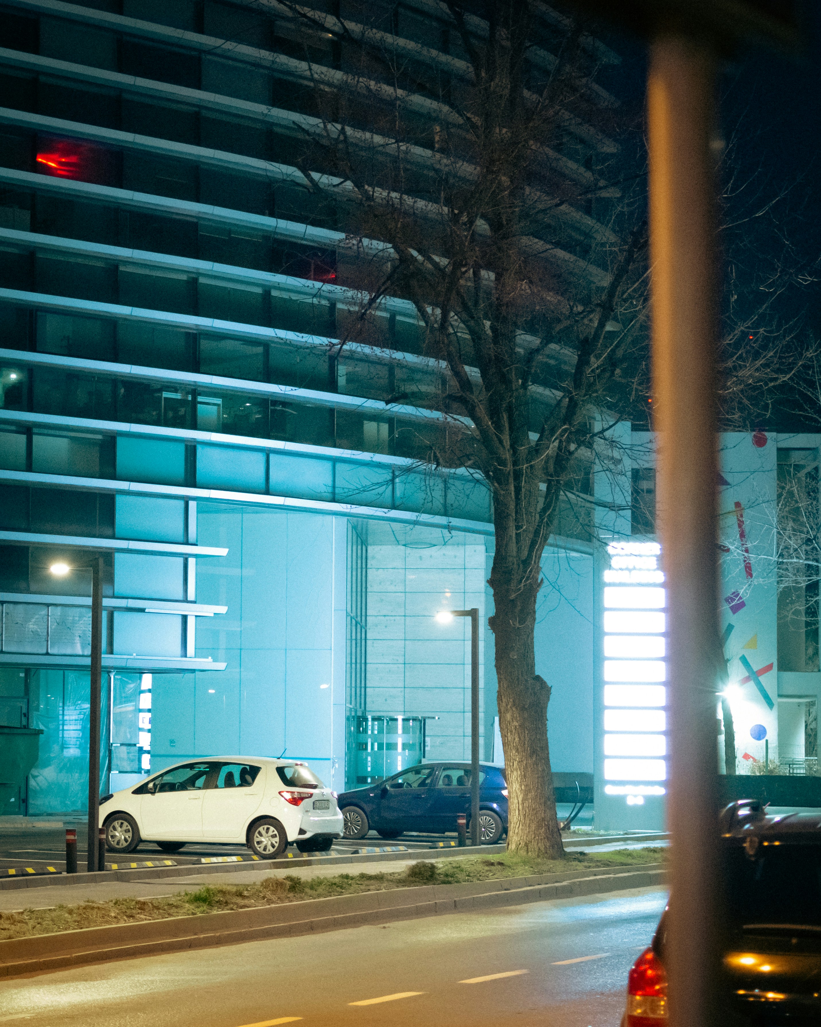 Blue Glass Lane: A White Hatchback Framed by Neon-Flooded Facades