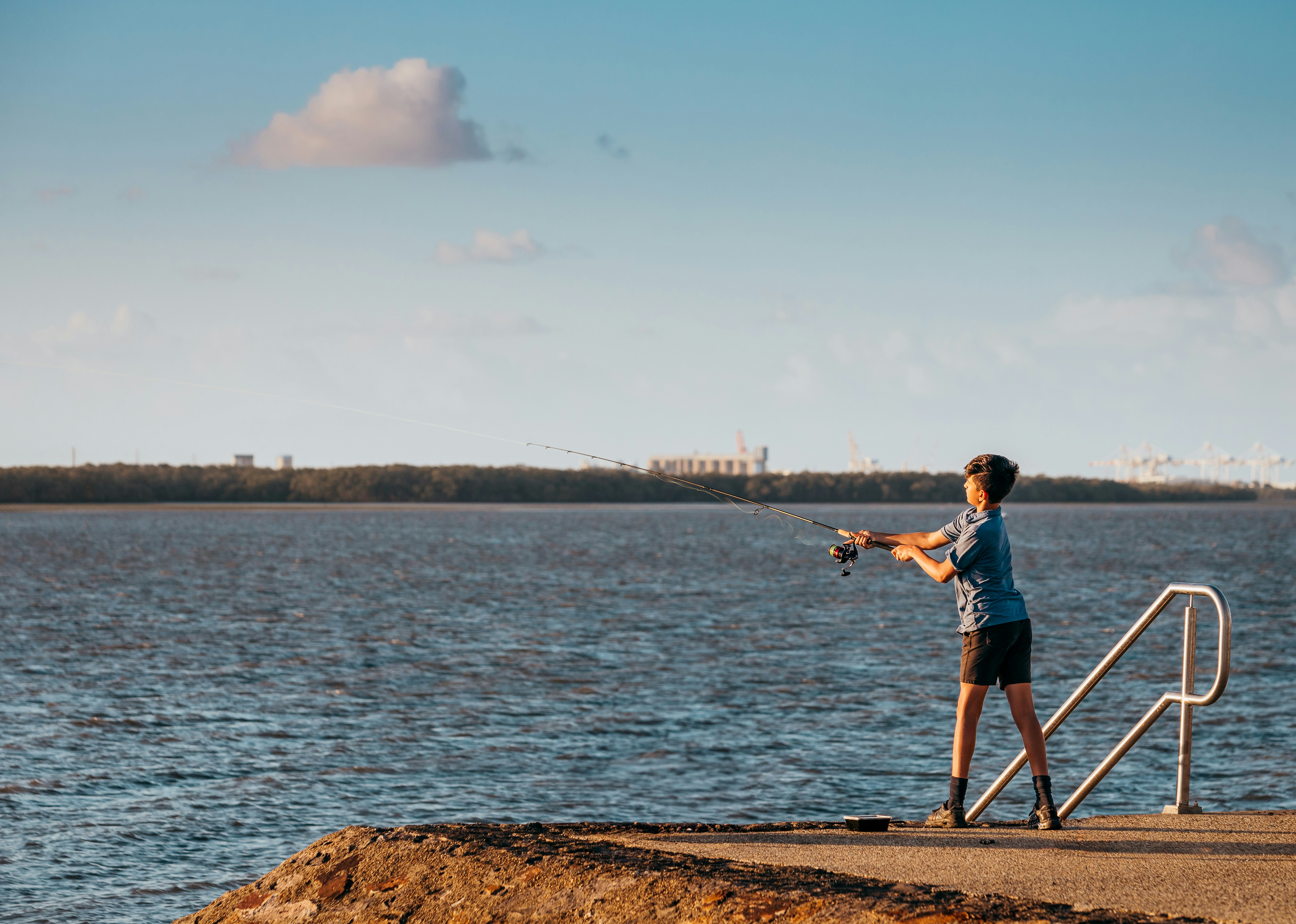 Young boy casting a fishing line from a rocky shore into a calm body of water under a clear sky.