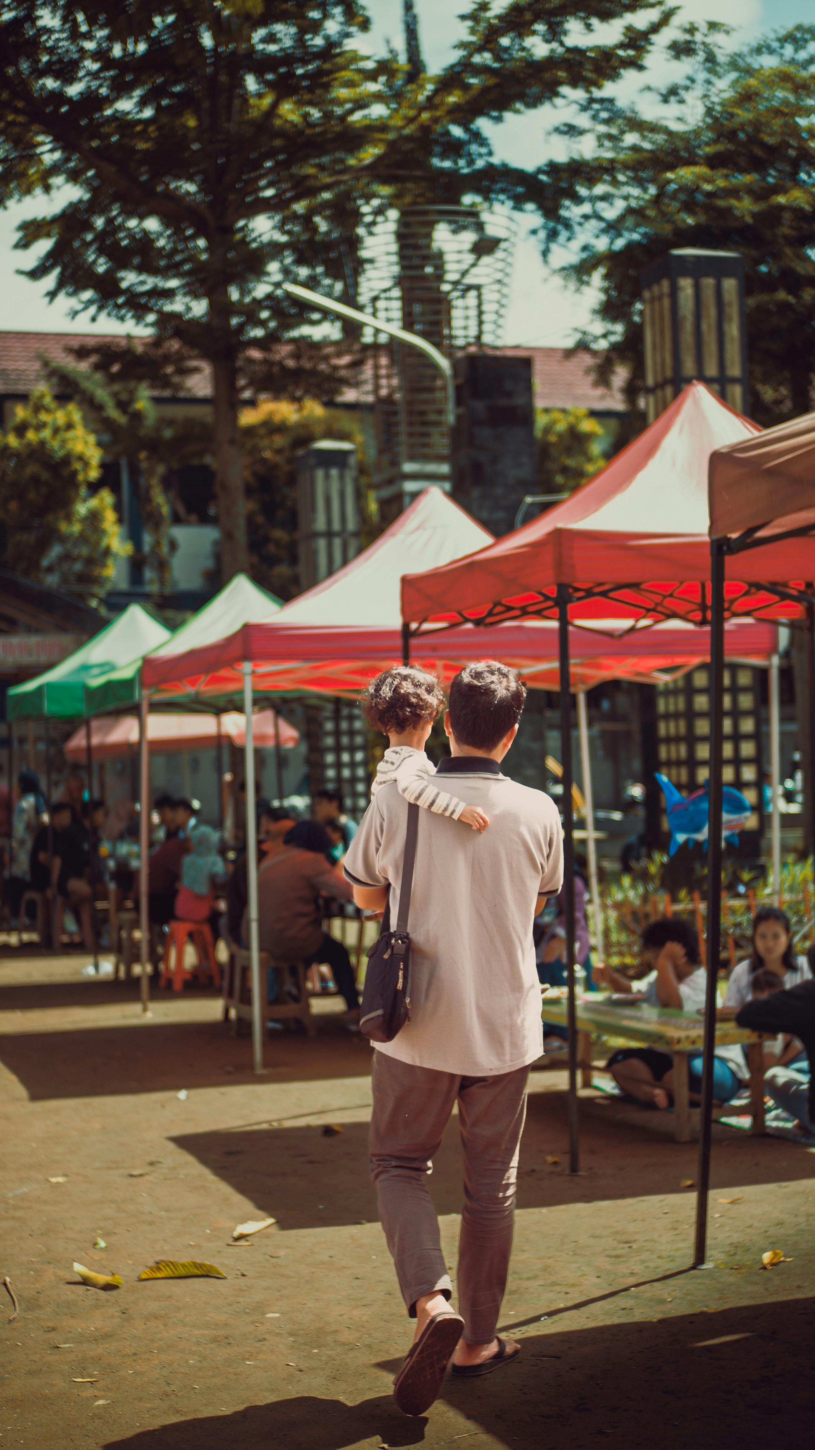 Sunlit Market Aisle: A Father Carries His Child Beneath Colorful Tents