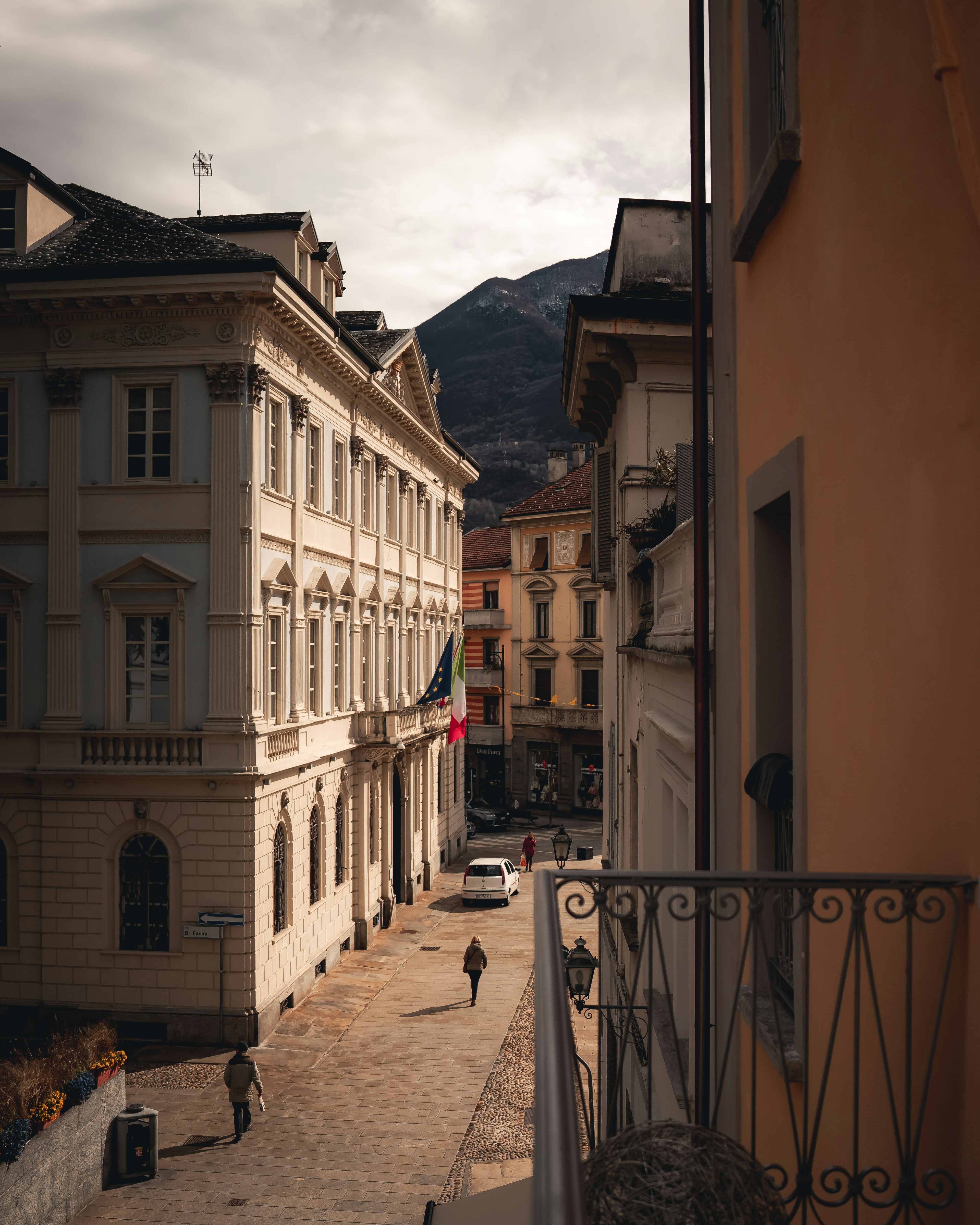 Sunlit town hall in Domodossola with mountain backdrop and a lone pedestrian on the cobblestone street.