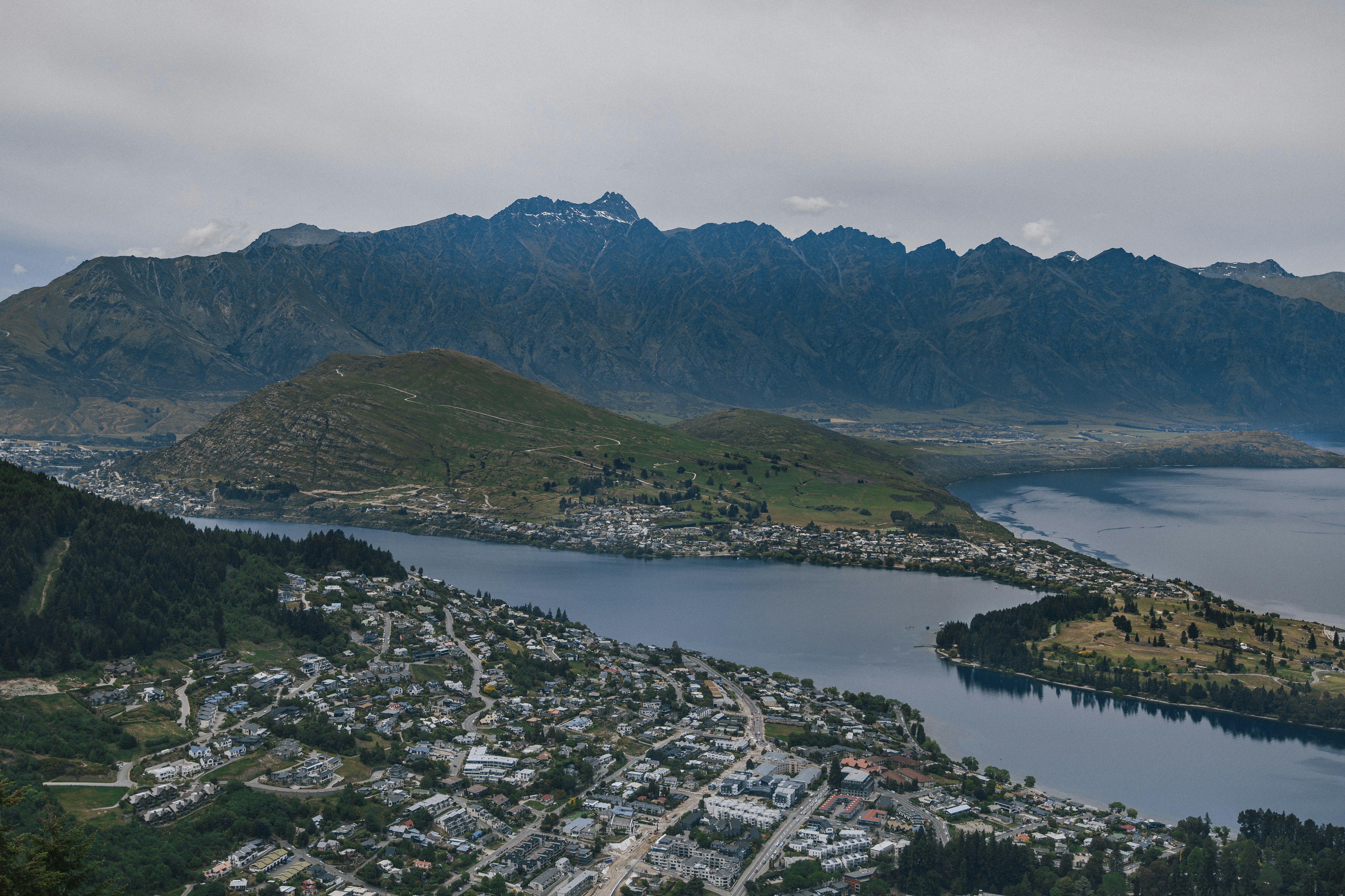 This breathtaking image captures a sweeping view of a picturesque lakeside town nestled beneath towering, rugged mountains. The scene is composed with a harmonious blend of deep blues, lush greens, and muted earth tones, enhanced by soft, diffused lighting that creates a tranquil atmosphere. The striking contrast between the vibrant town below and the dramatic mountain range above makes this landscape visually captivating.