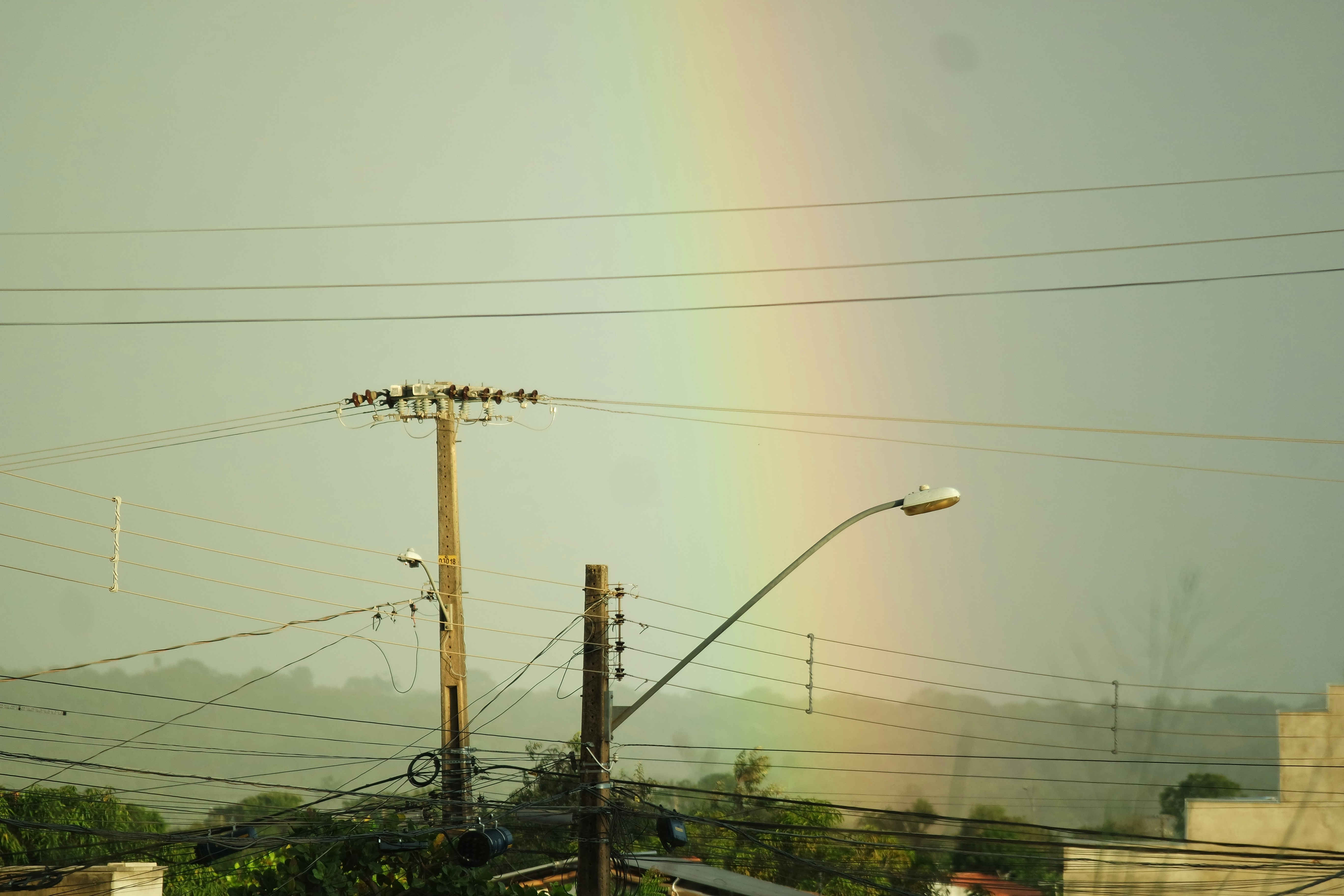 A photograph capturing a rainbow arching over a suburban landscape, framed by power lines and a streetlamp. The scene juxtaposes man-made infrastructure with a natural arc.