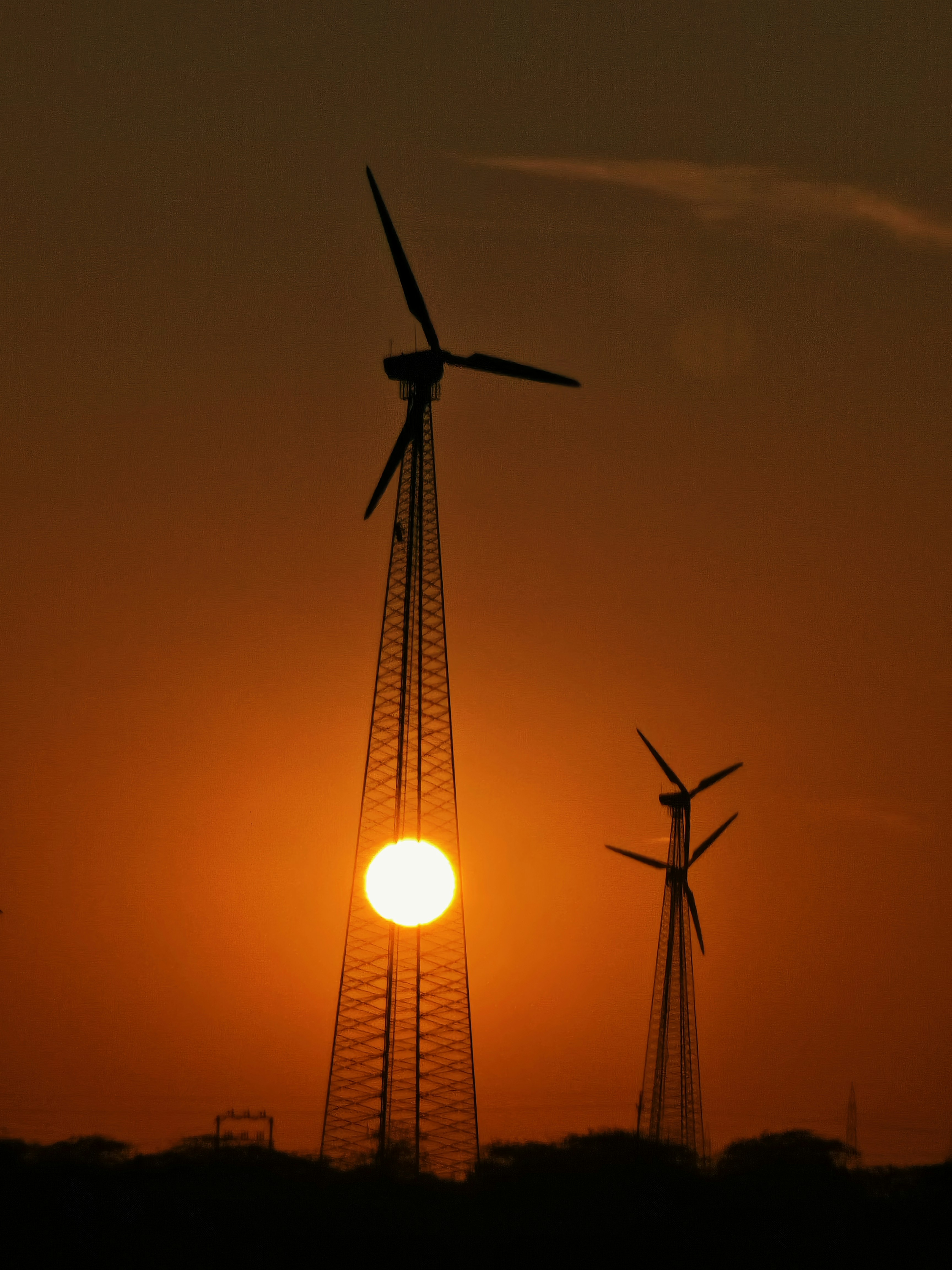 Two wind turbines stand as silhouettes against a fiery sunset. The sun sits between the towers, creating warm backlight and a moody horizon.