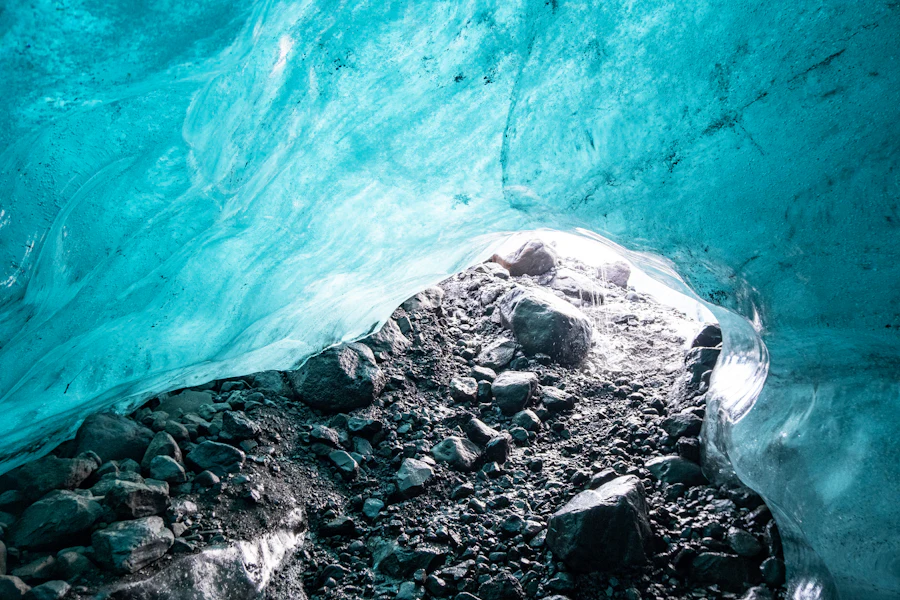 Inside a glowing turquoise ice cave, a rough rocky path leads toward a bright opening at the far end. Curved ice walls frame the entrance with cool light reflecting across the chamber.