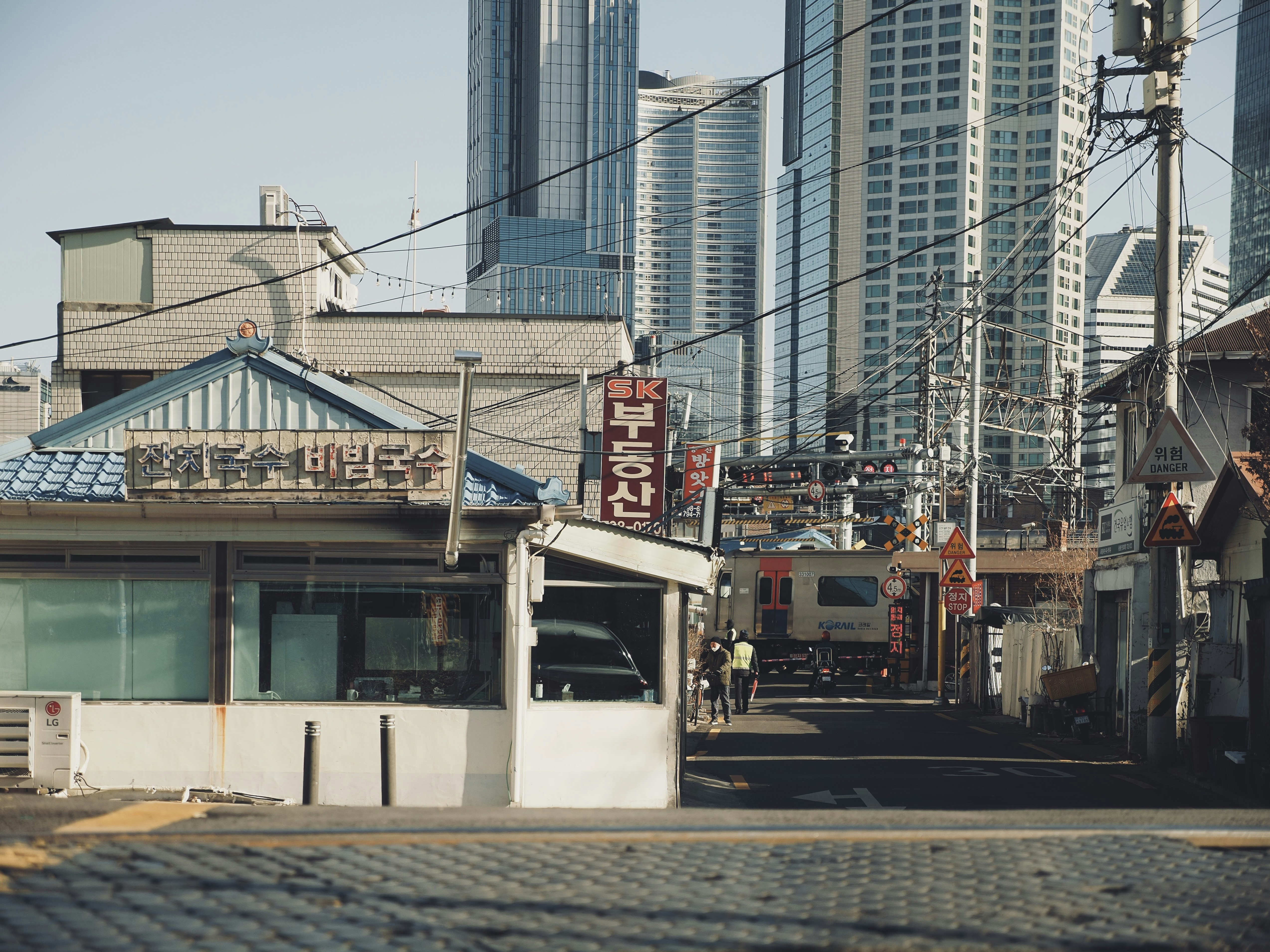 A street scene in a Korean city with low-rise shops in the foreground and modern high-rise towers in the distance.