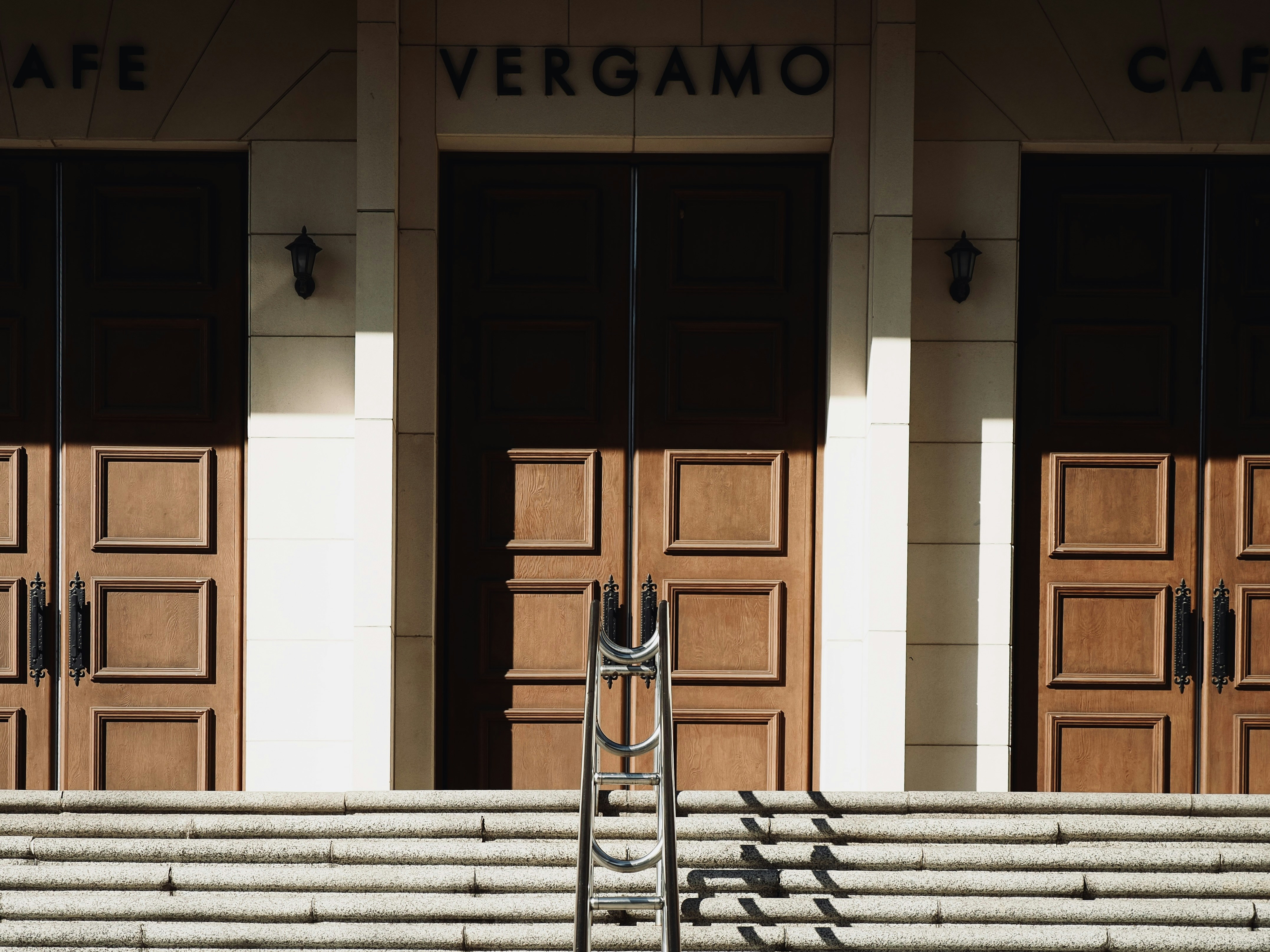 Photograph of a sunlit façade with three wooden doors, stone steps, and a metal railing in front.