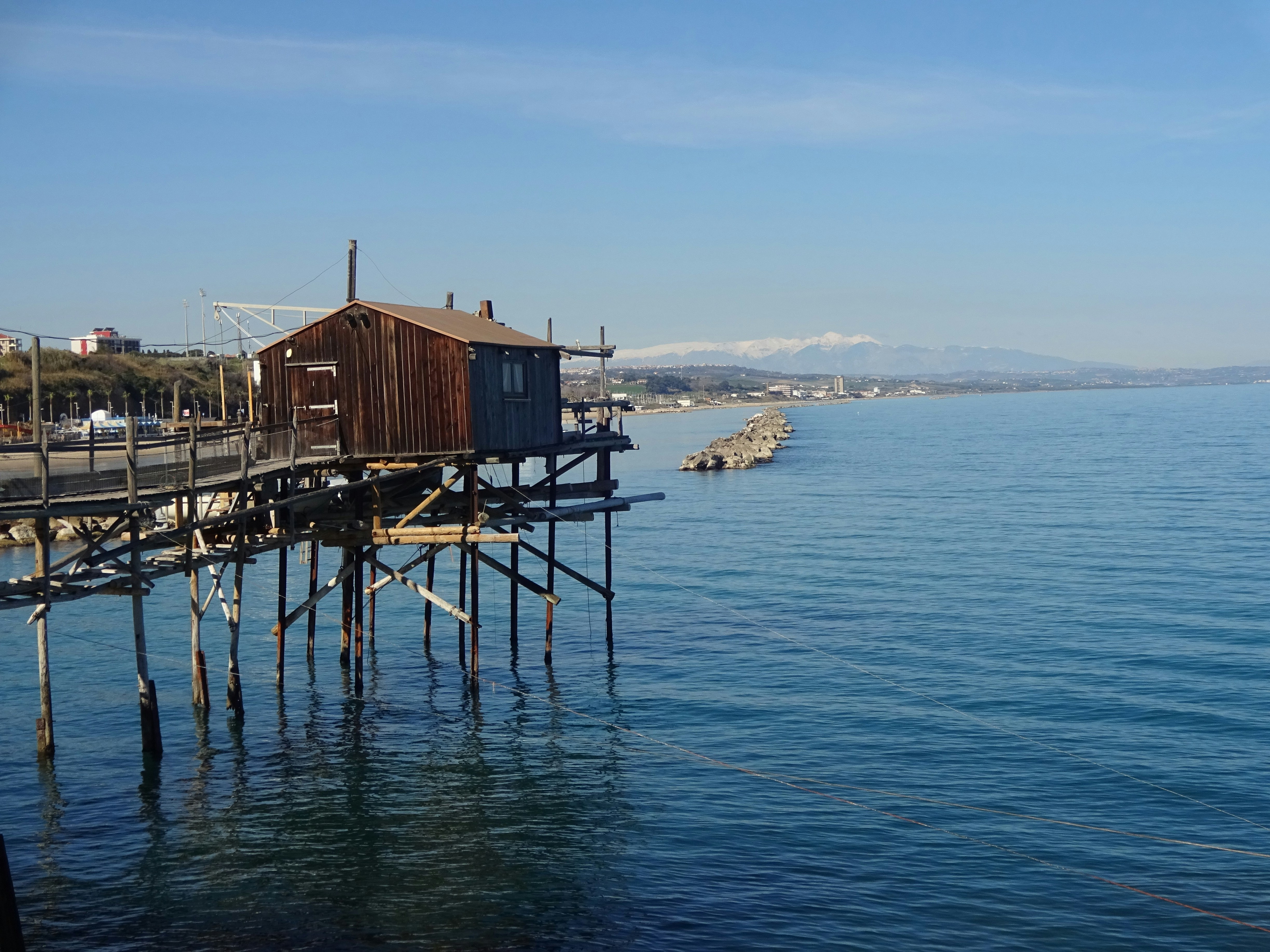 Wooden dock with a small house extending over calm blue sea beneath a clear sky.