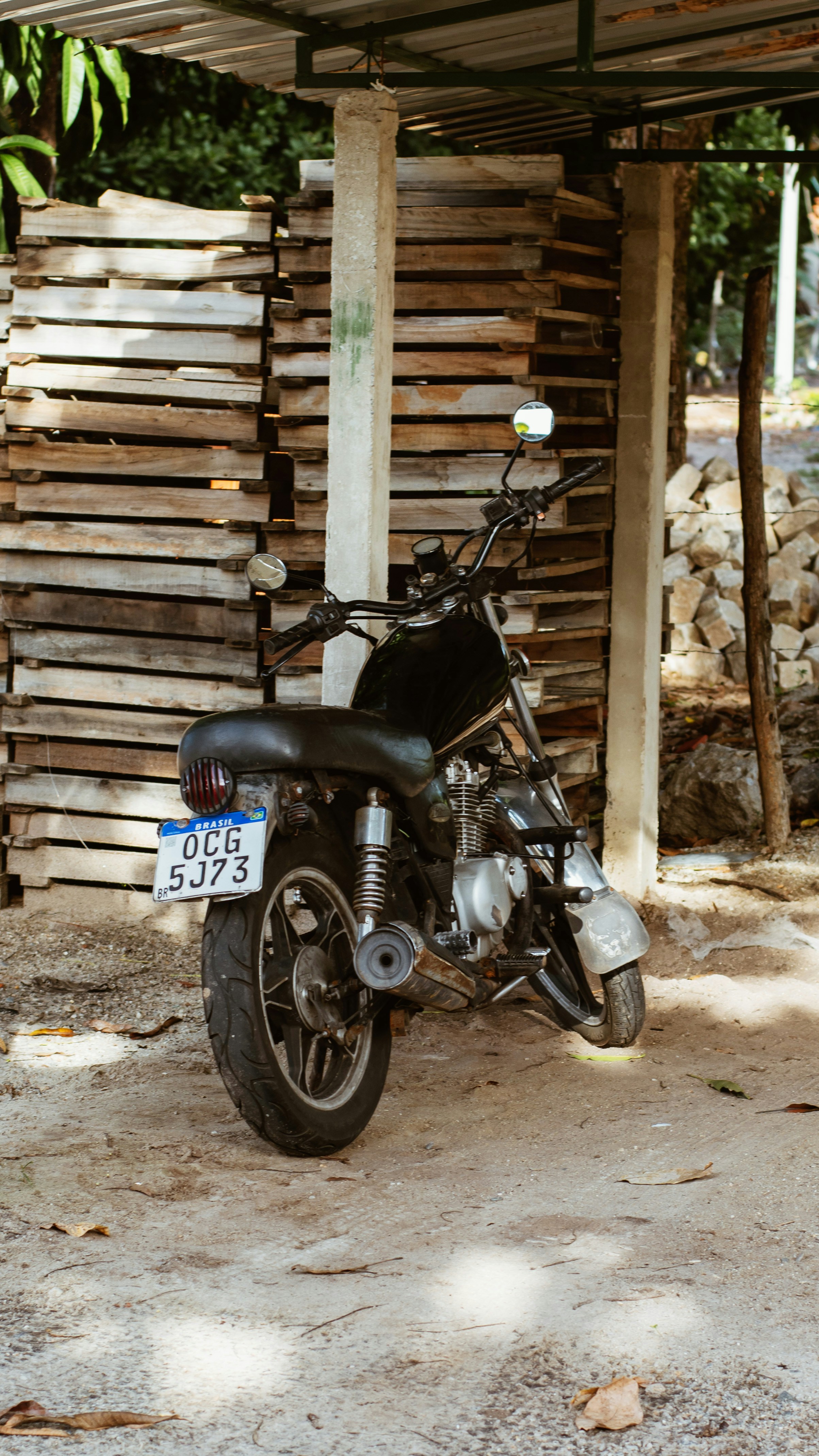 Shadowed Steel: A Retro Motorcycle Paused Among Weathered Wood Piles