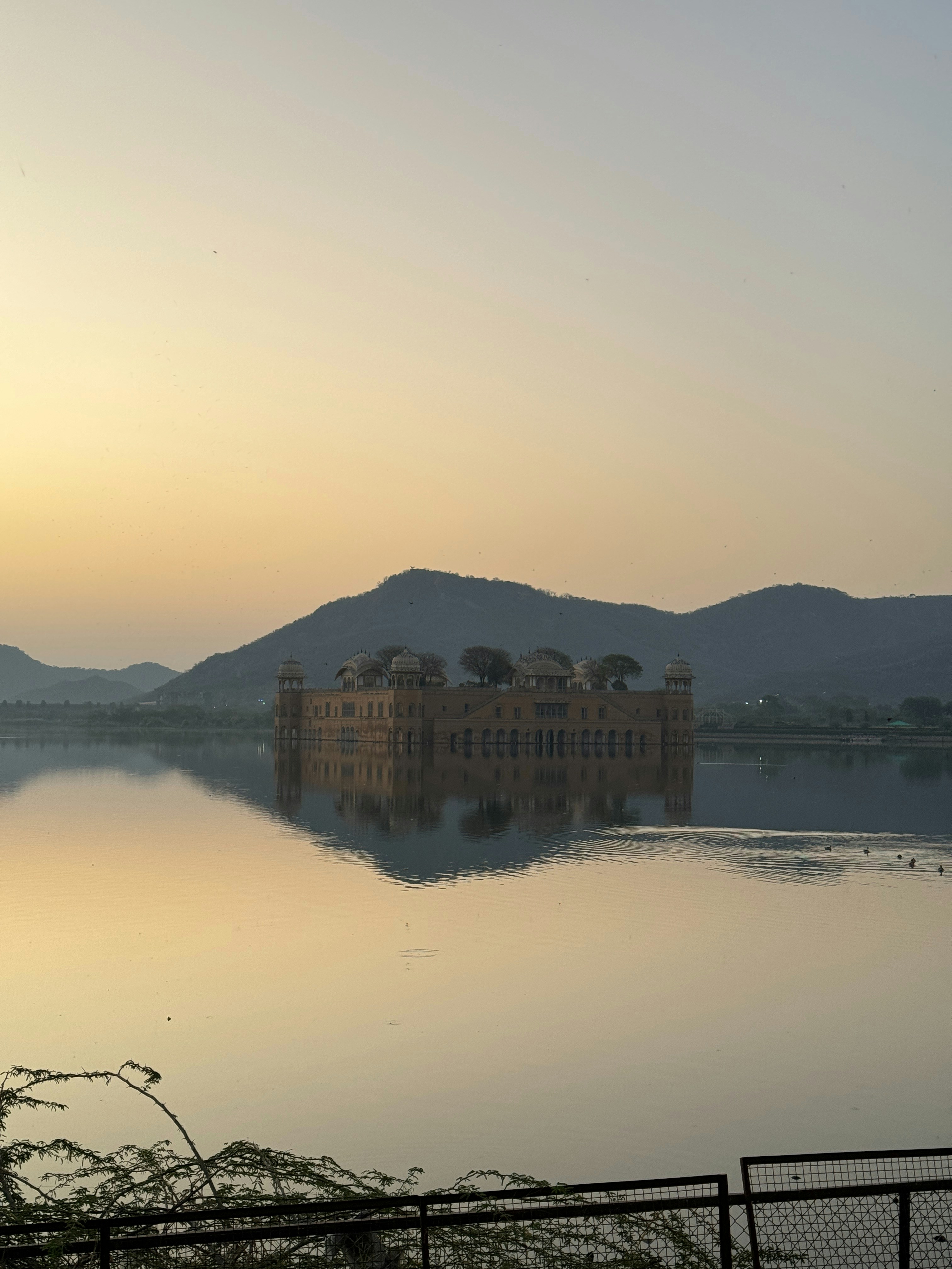 Jal Mahal palace reflecting in the tranquil waters of a lake at sunrise with distant mountains silhouetted against the sky.