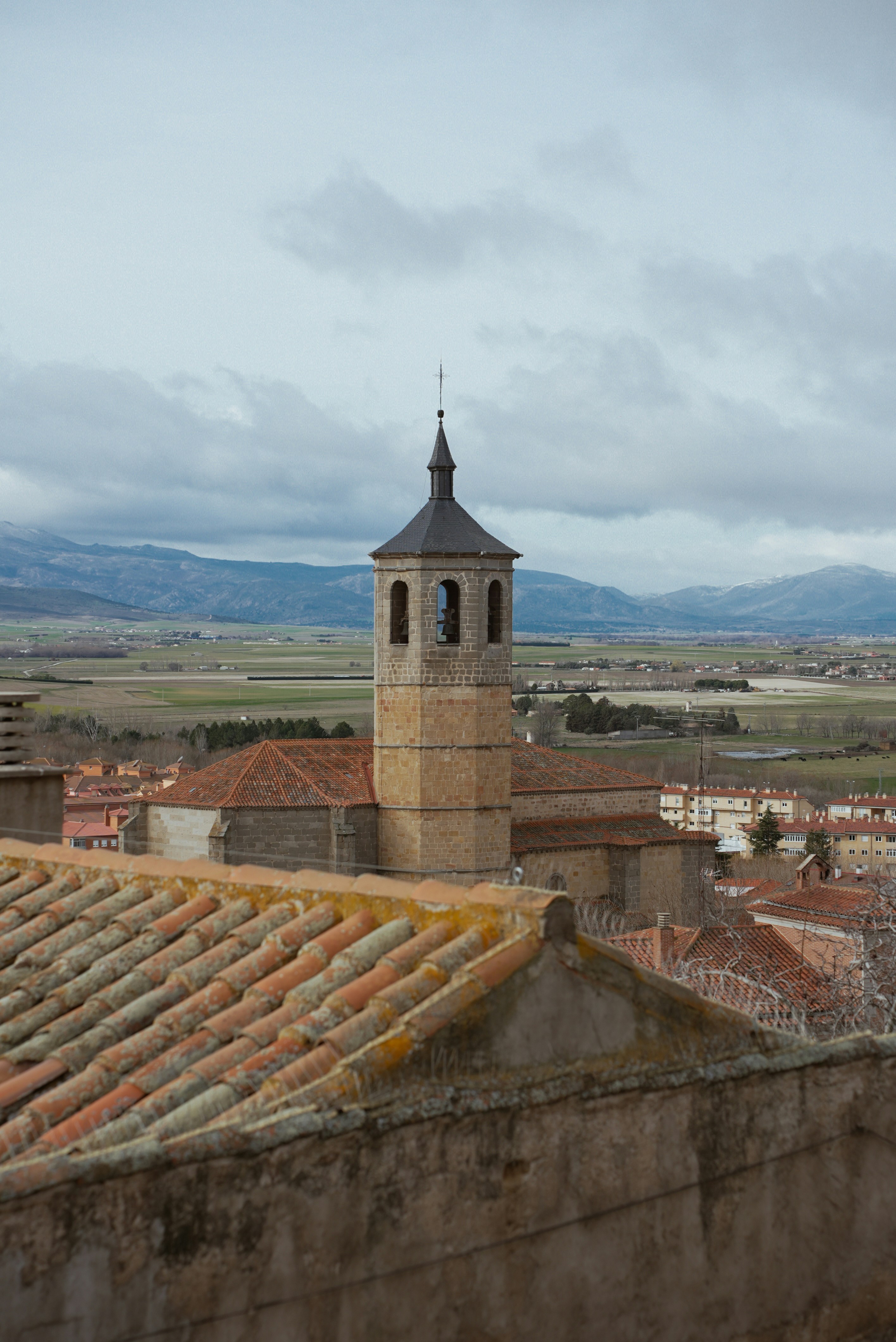 Stone bell tower rises above red-tiled roofs, with a flat plain and distant mountains under a cloudy sky. The composition emphasizes architectural detail and a tranquil townscape.