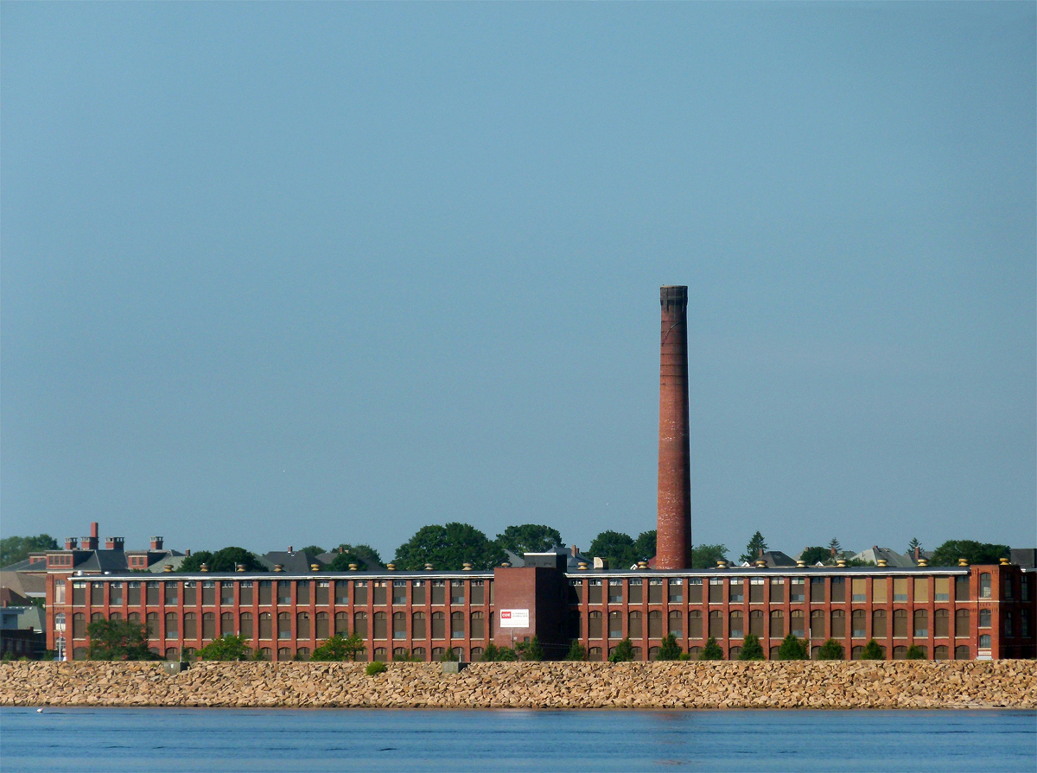 Large brick factory building with tall smokestack beside a calm body of water.