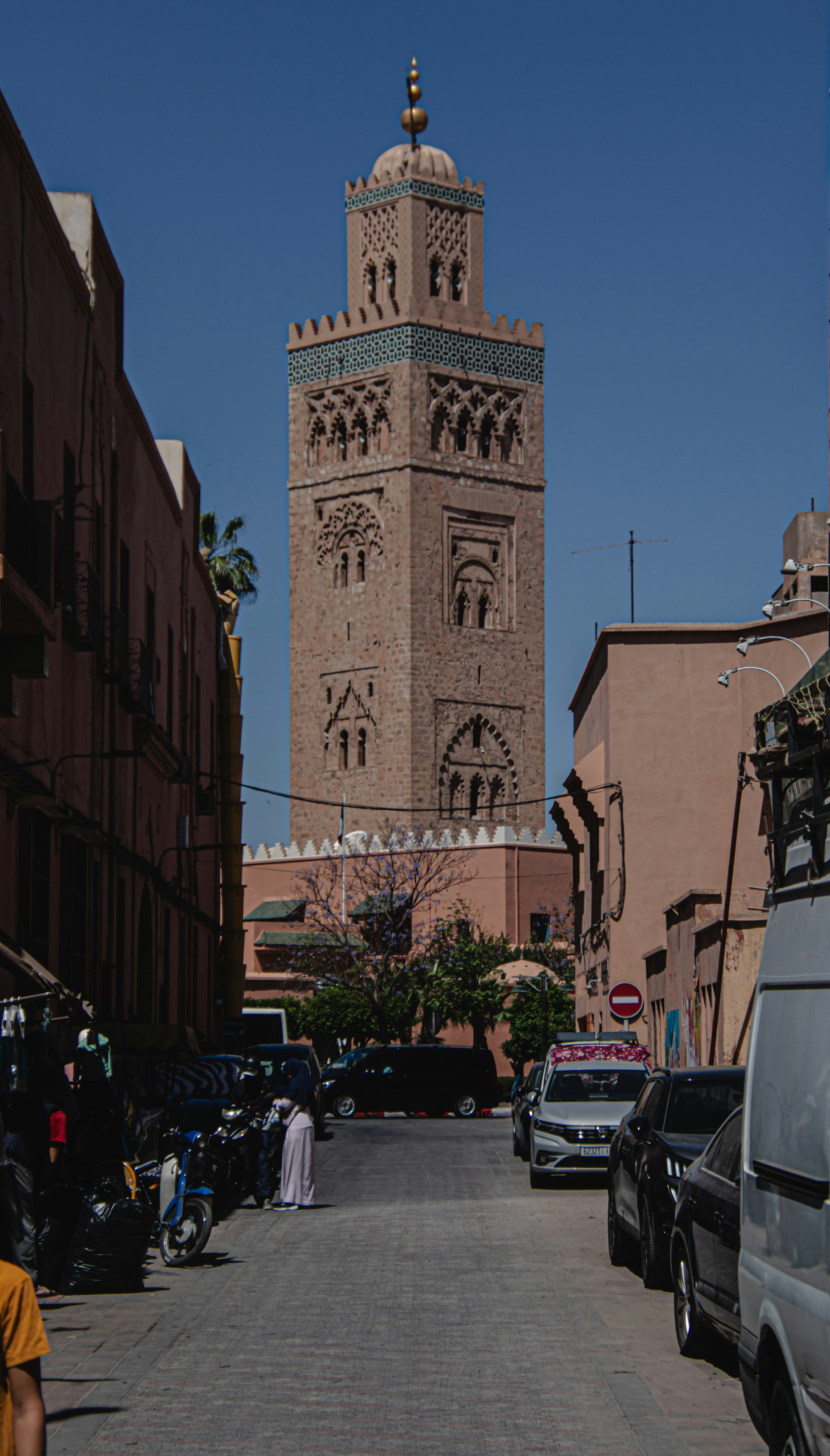 Sunlit Moroccan minaret rises above a narrow street flanked by pinkish buildings, with parked cars and pedestrians in the foreground.