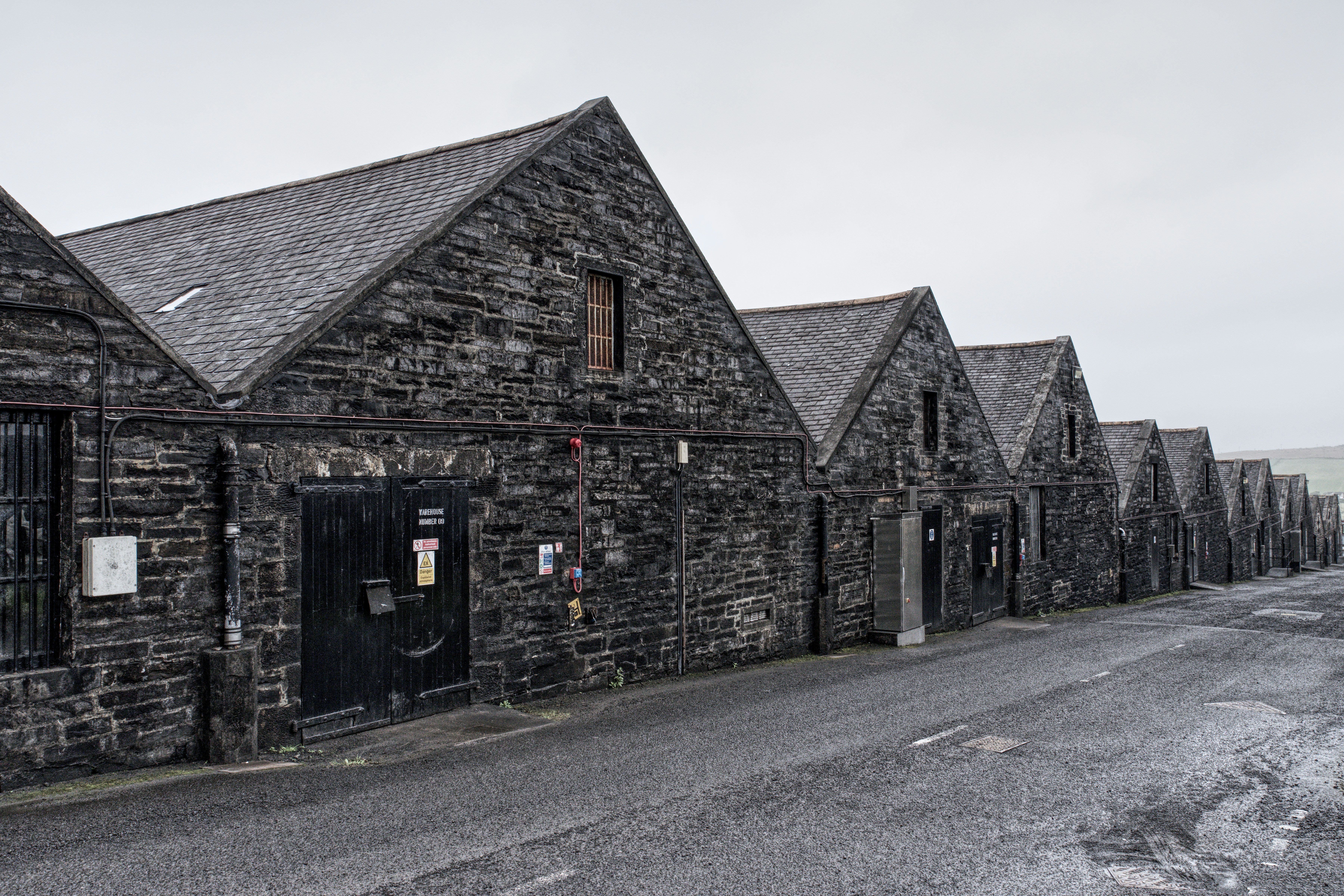 Row of rustic stone buildings under a gray sky, with a wet road leading into the distance.