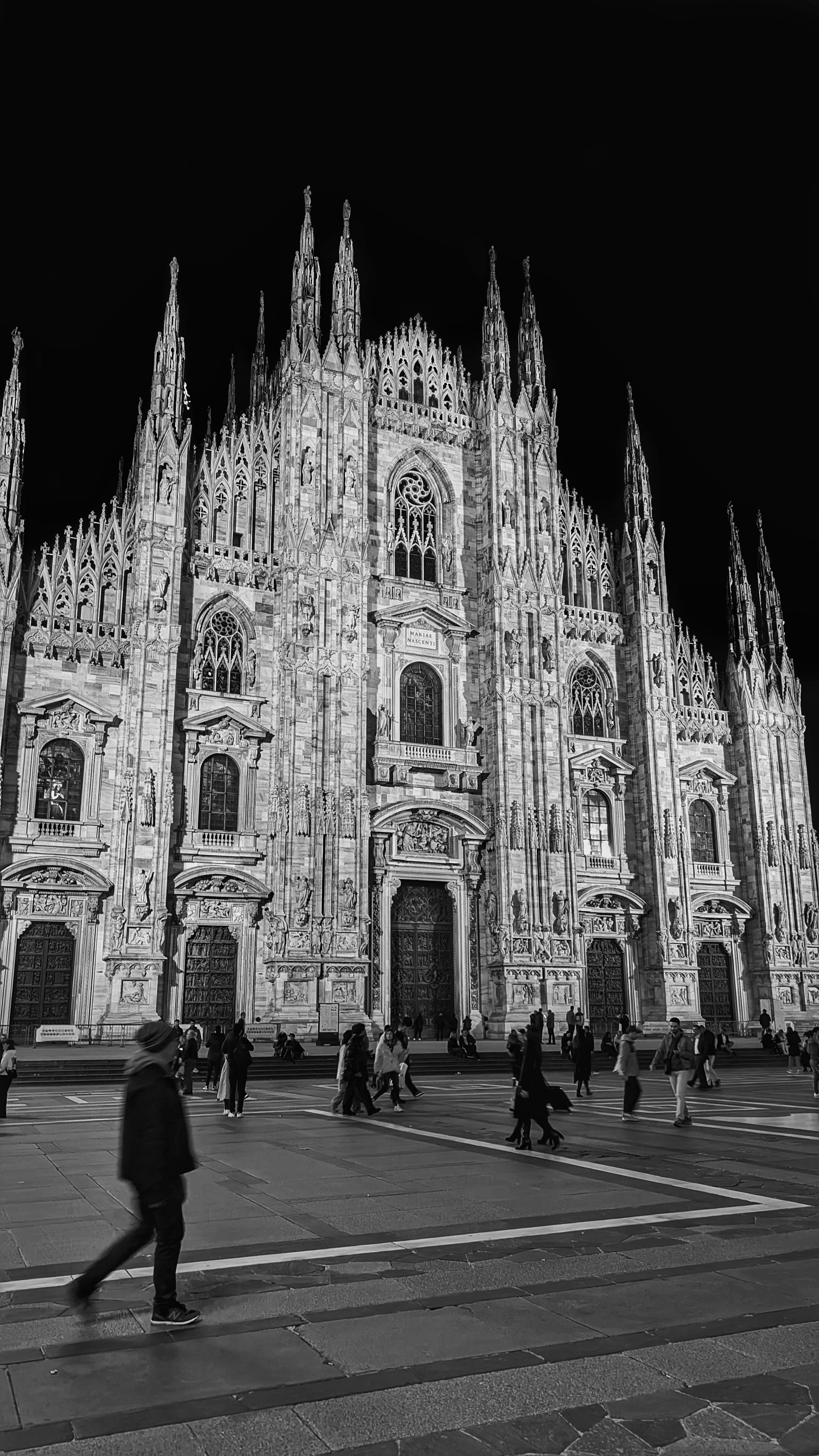 Gothic cathedral facade bathed in bright illumination against a dark sky. Pedestrians traverse the broad plaza in the foreground.