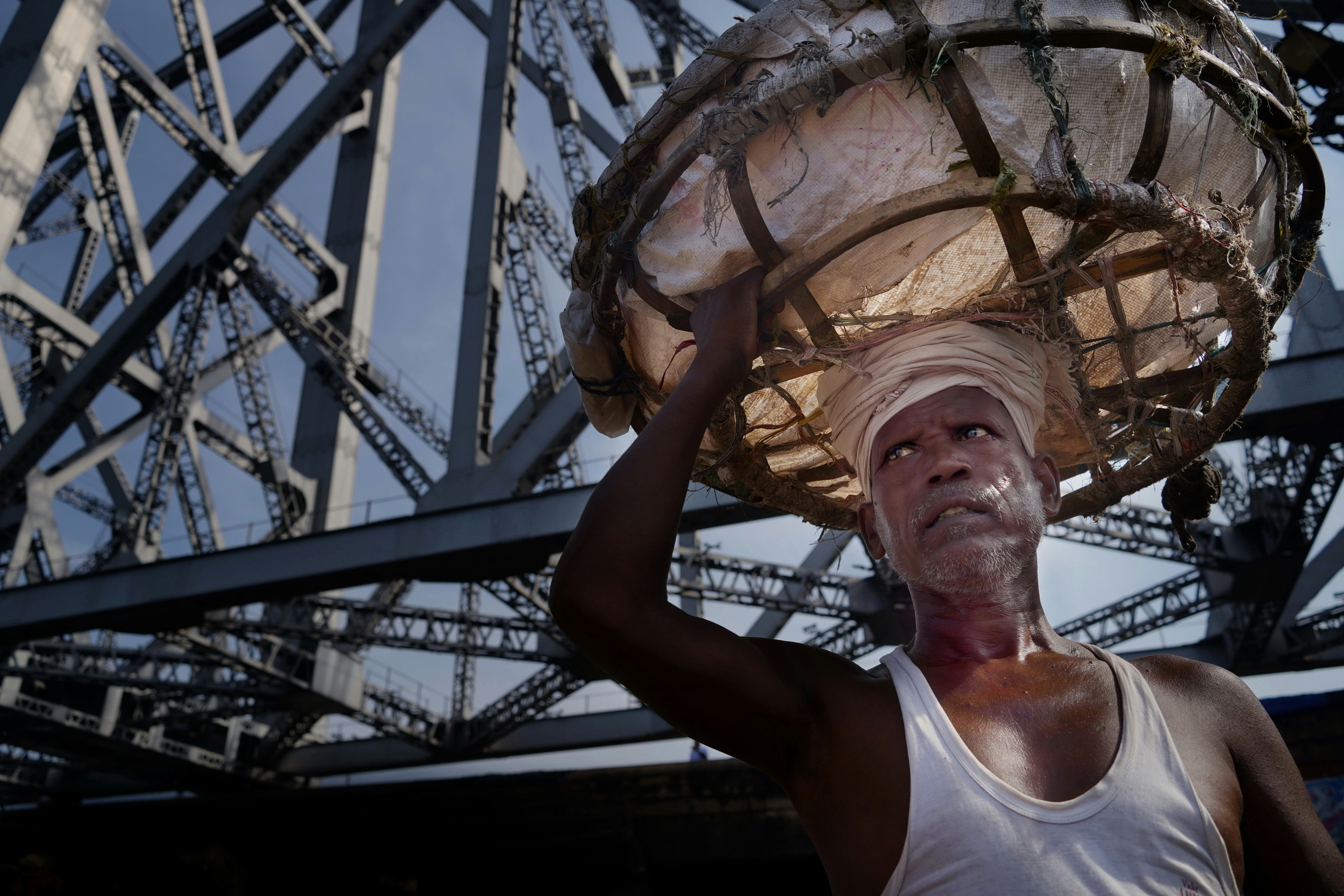 Man carrying a large basket under a towering bridge structure.