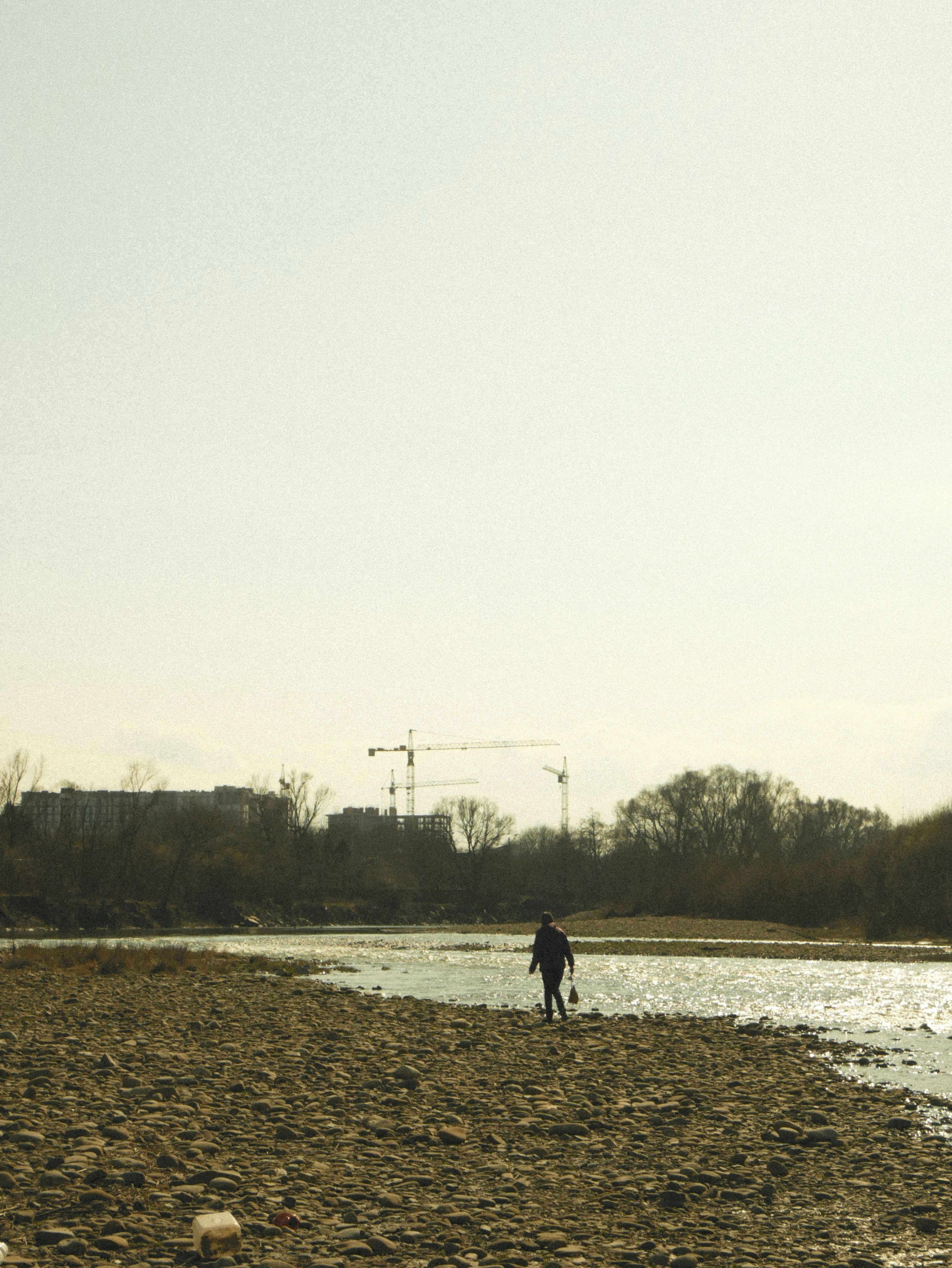 Pebbled Riverbank Stroll Beneath a Quiet Construction Horizon