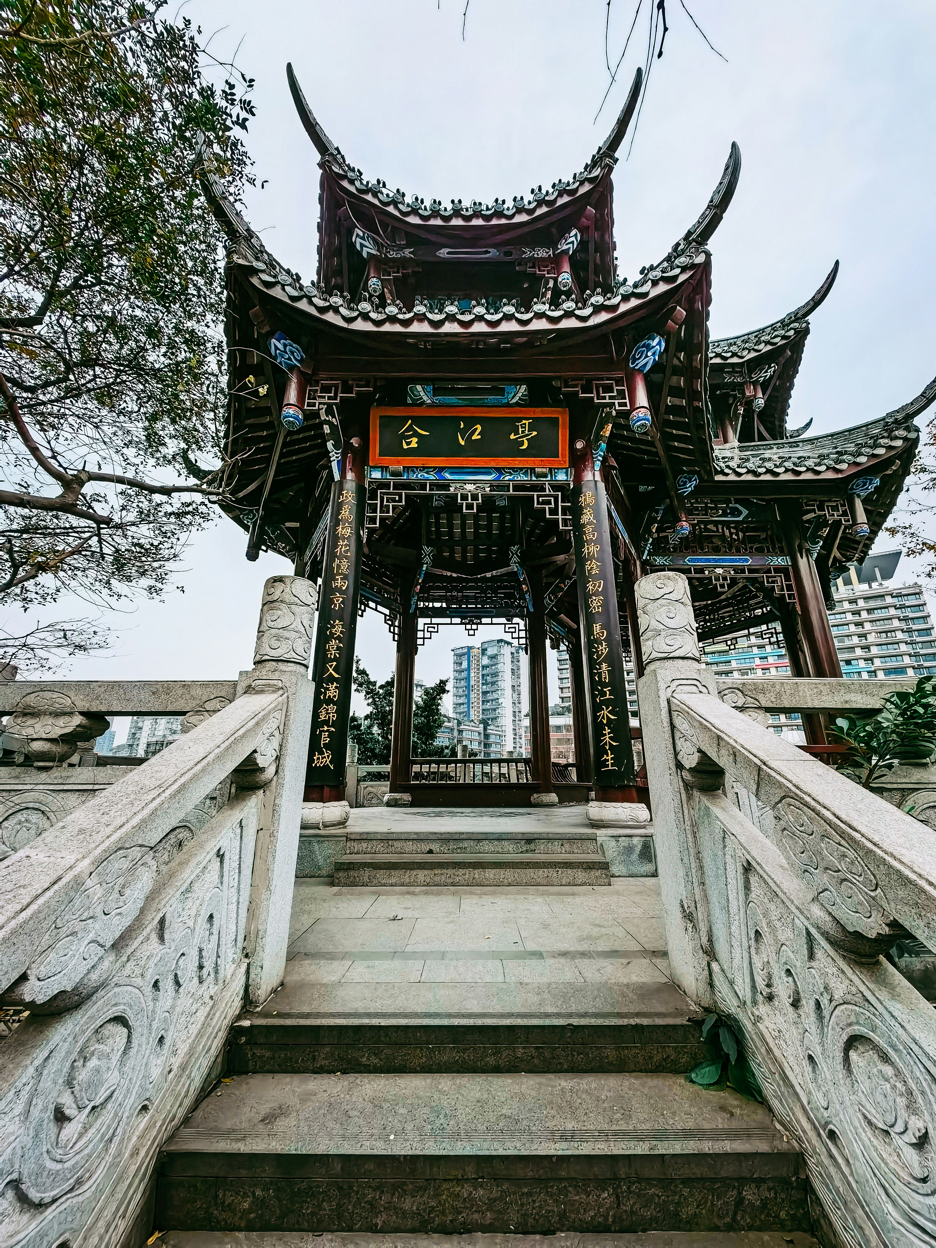 Low-angle photograph of an ornate Chinese pavilion with curved eaves and carved railings, rising above stone steps. The urban skyline in the background contrasts with the traditional architecture.