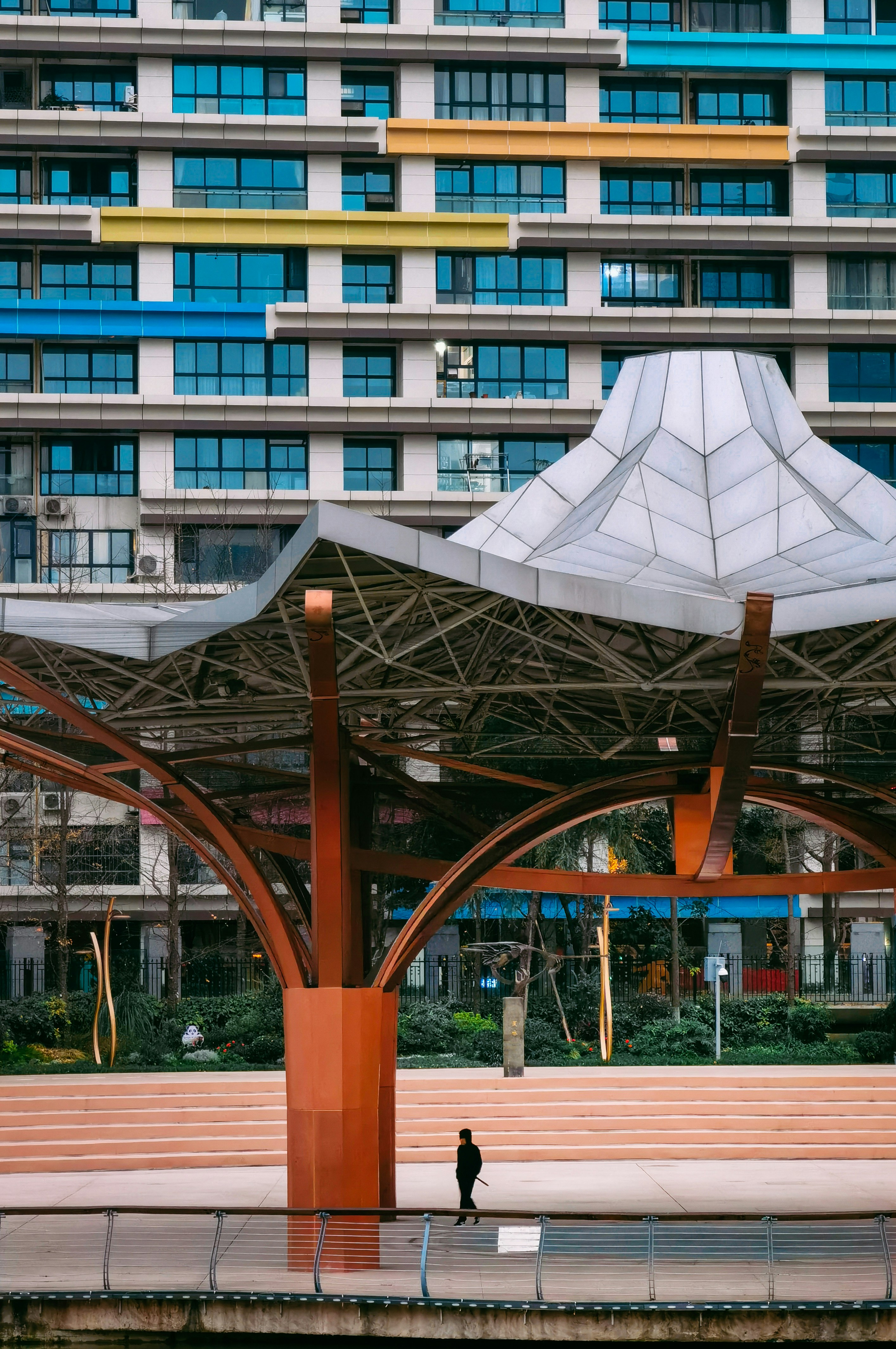A photograph of a modern urban scene with a striped building backdrop, an orange arched canopy, and a solitary figure along the walkway.