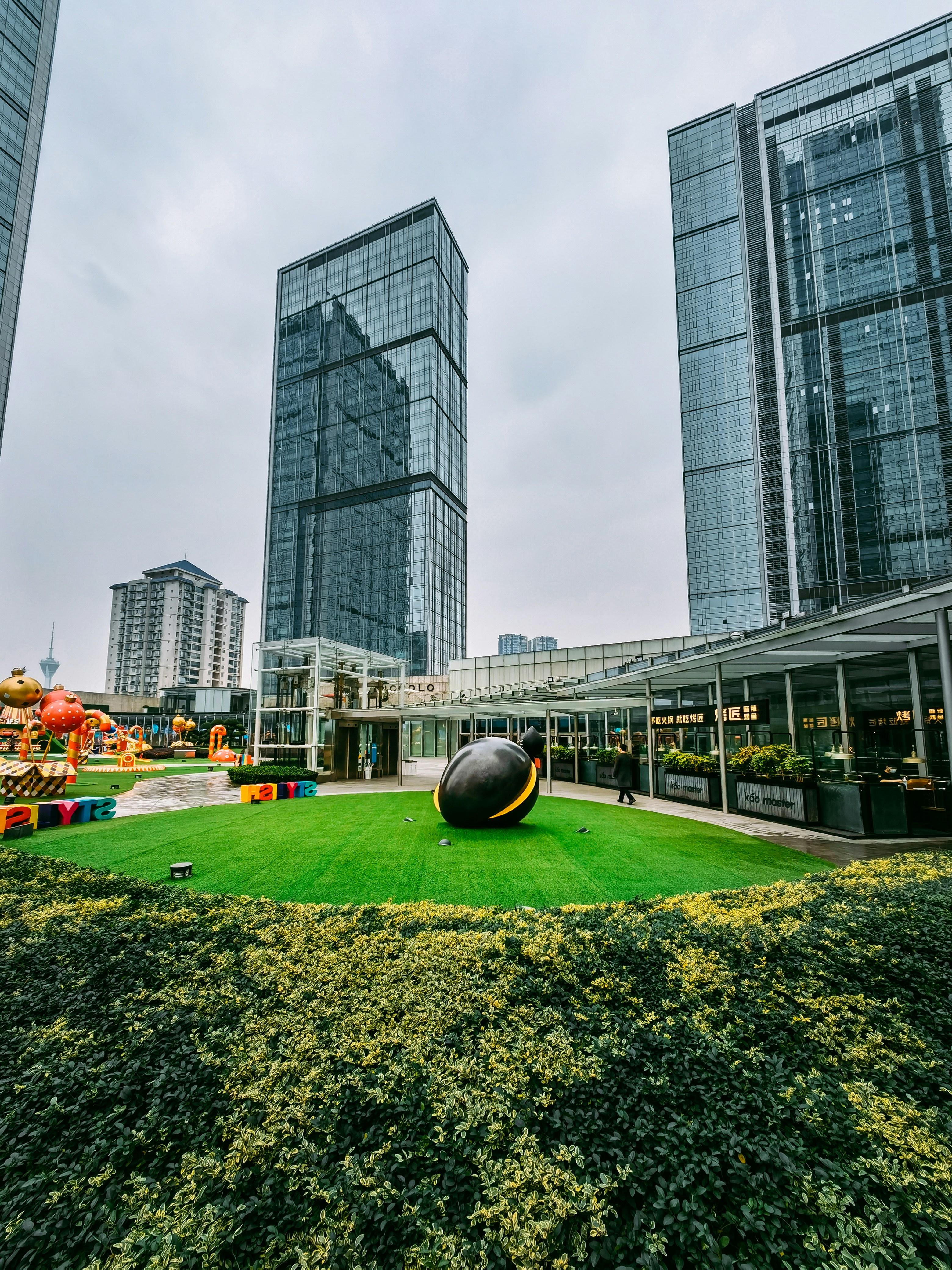 Photograph of a city plaza framed by towering glass skyscrapers, featuring a black spherical sculpture on a bright green lawn and a hedge border in the foreground.