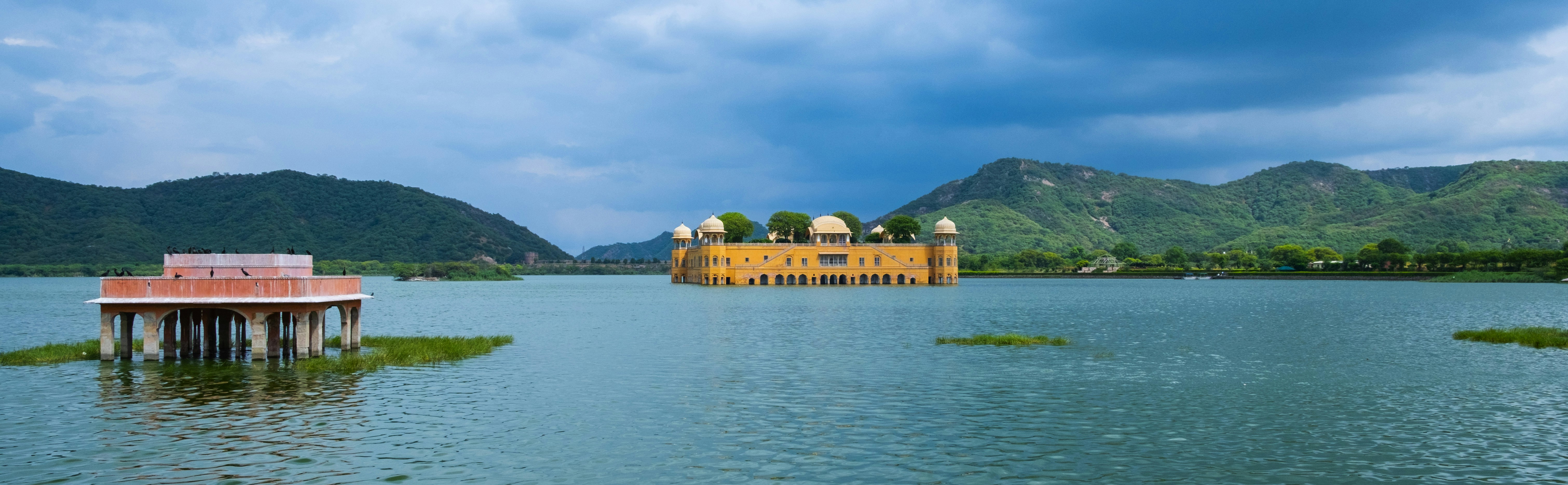 Palace surrounded by calm lake with mountains in the background under a cloudy sky.