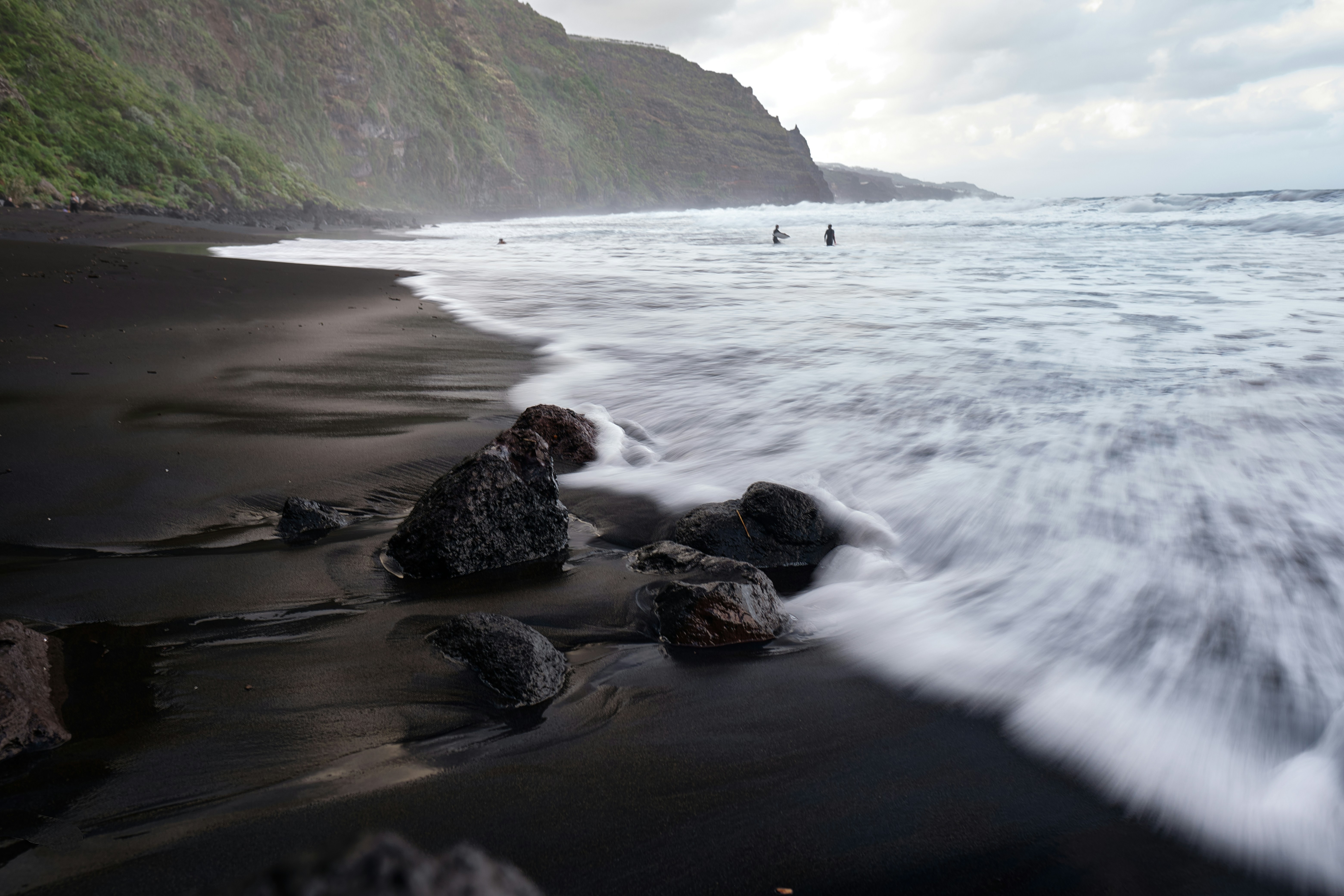 Waves gently lap against dark volcanic sand at a secluded beach under a cloudy sky.
