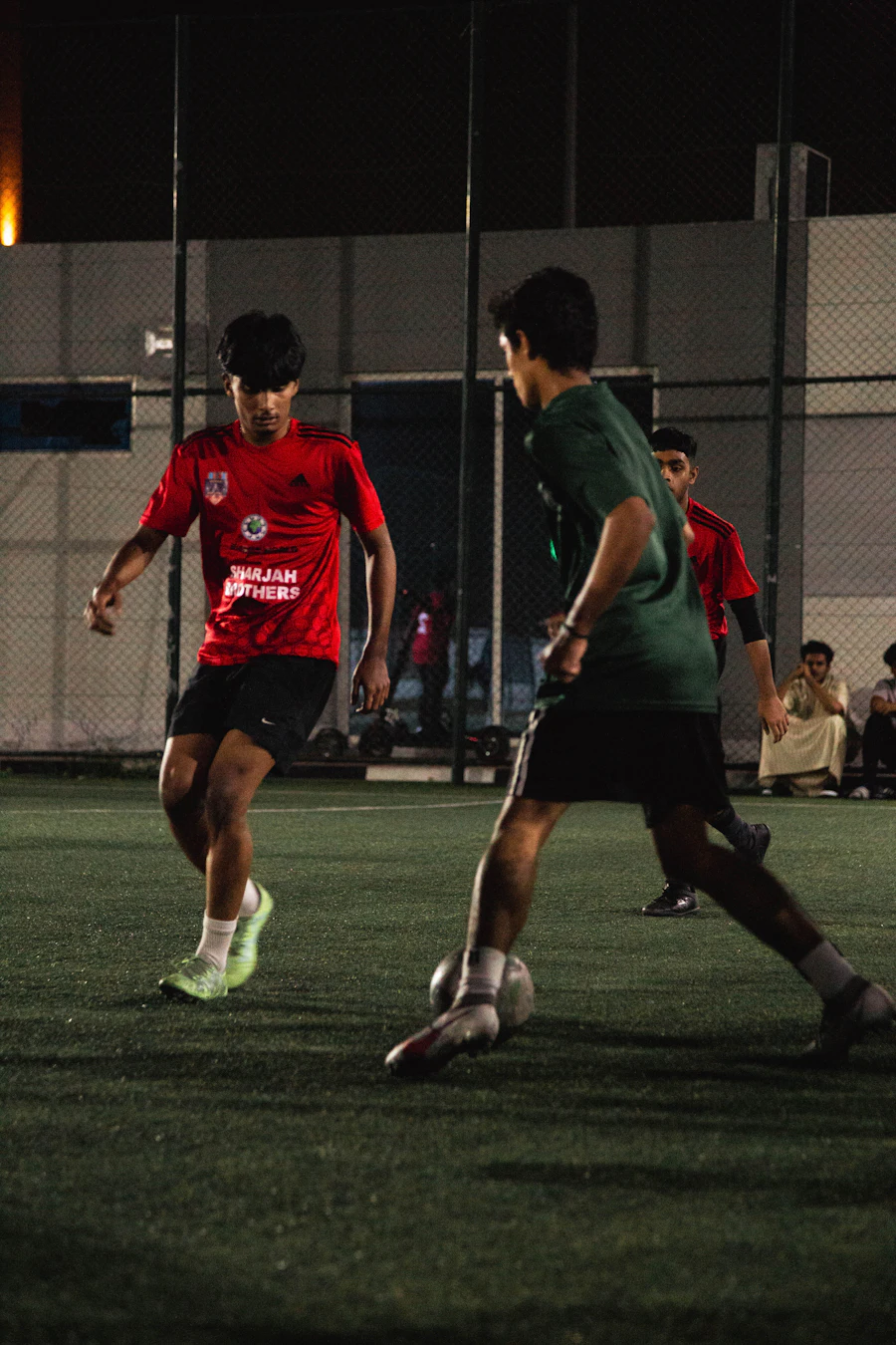 Two young players contest a dribbling pass on a floodlit artificial turf pitch at night.