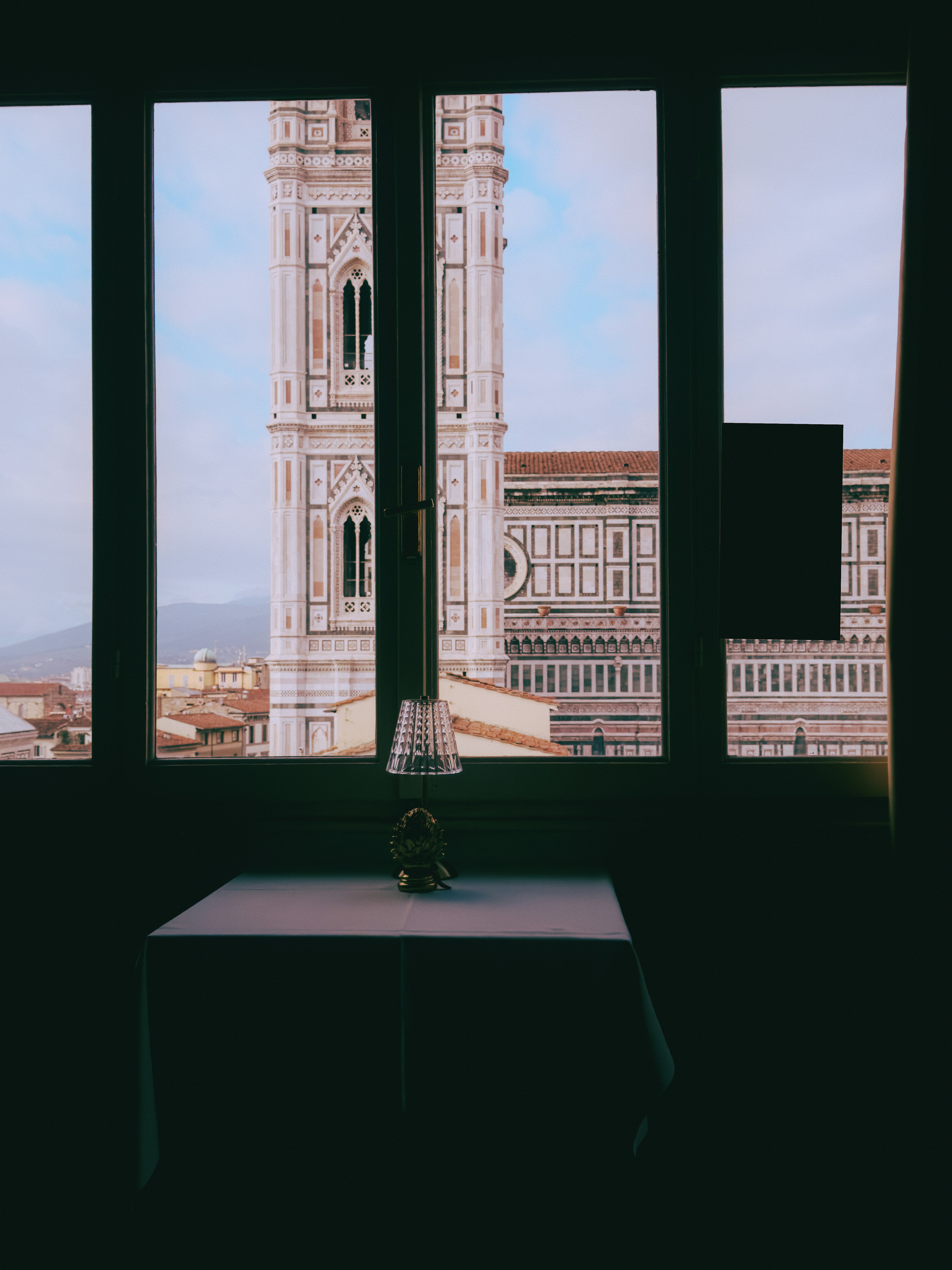 Dim interior with a lamp atop a white-draped table, framed by window panes revealing Florence’s Giotto's Campanile.