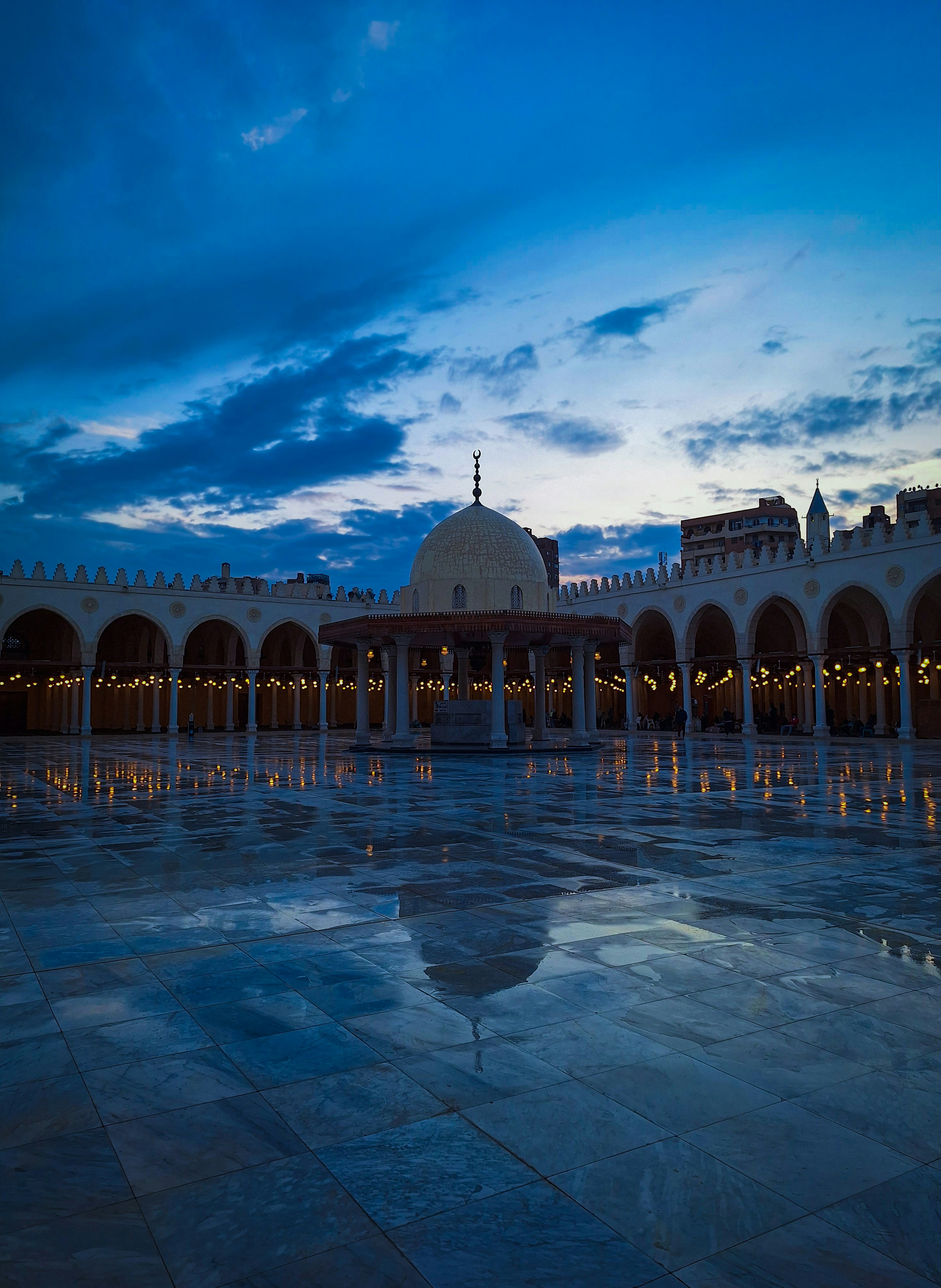 Blue-hour courtyard around a domed mosque with an arcade of arches, illuminated lamps, and wet marble reflecting the twilight sky.