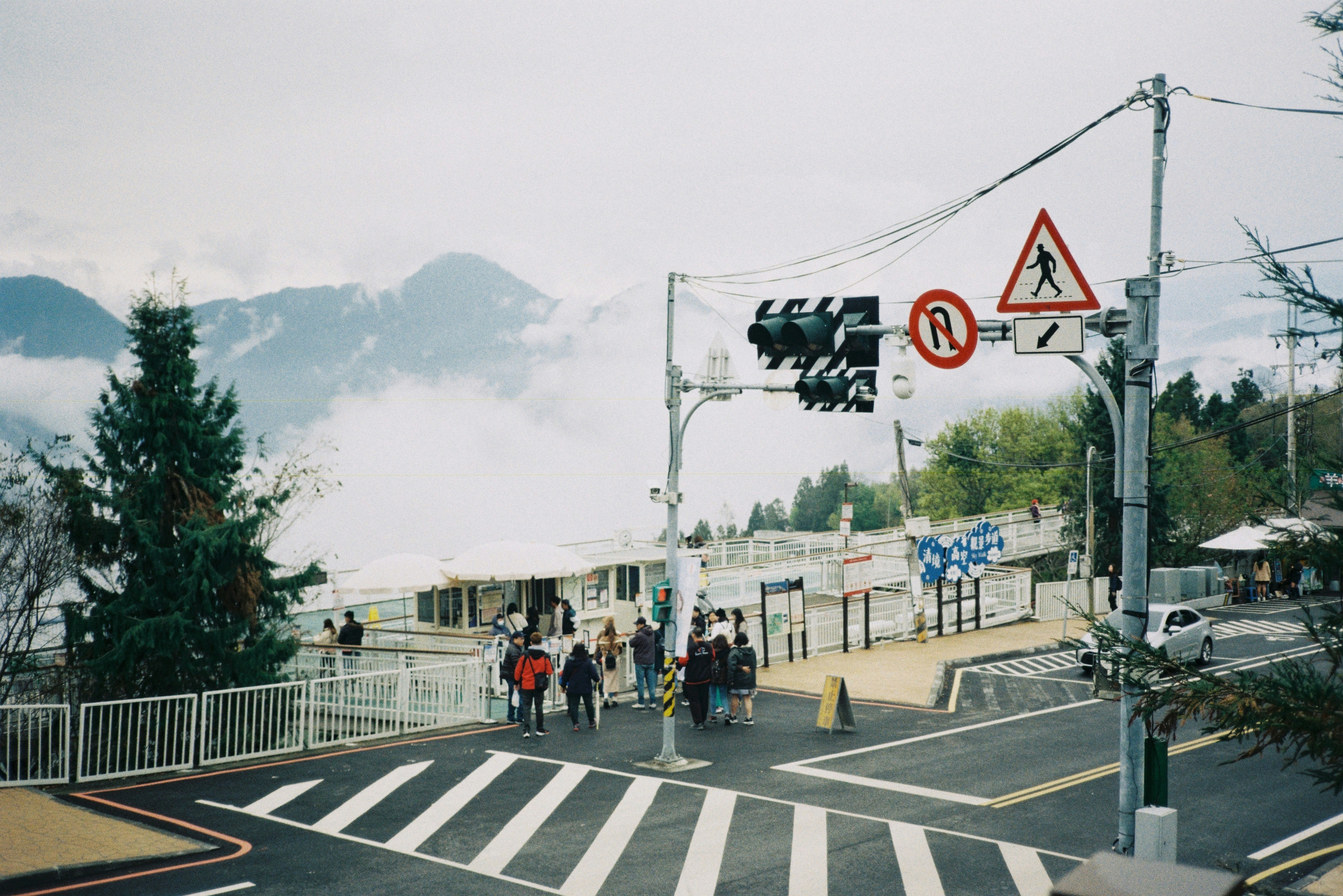 Pedestrians cross a street with misty mountains in the background on a cloudy day.