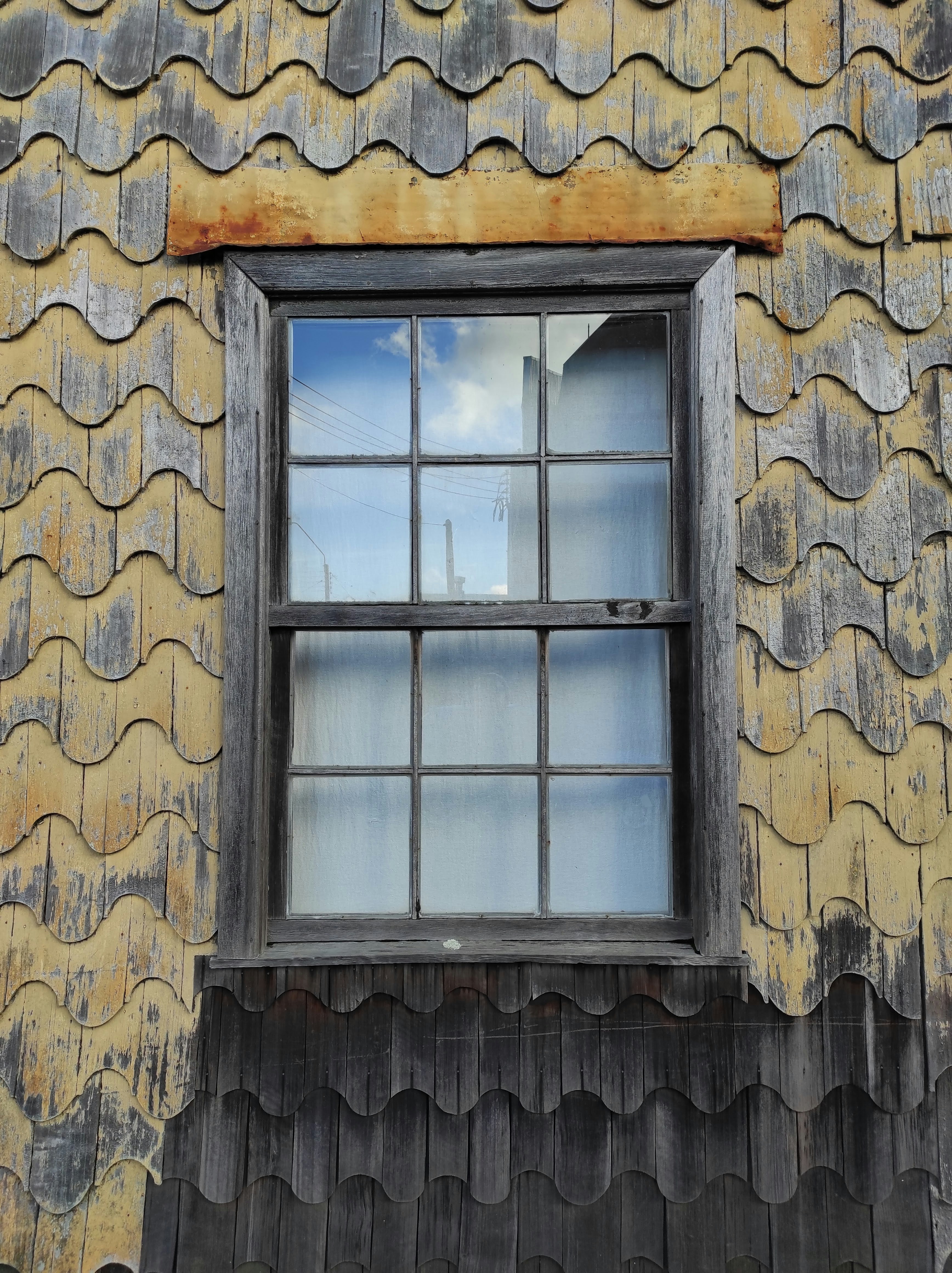 A weathered wooden window sits center-stage among yellow, wavy ceramic wall tiles, with a rusted trim casting an aged frame and reflections in the glass.