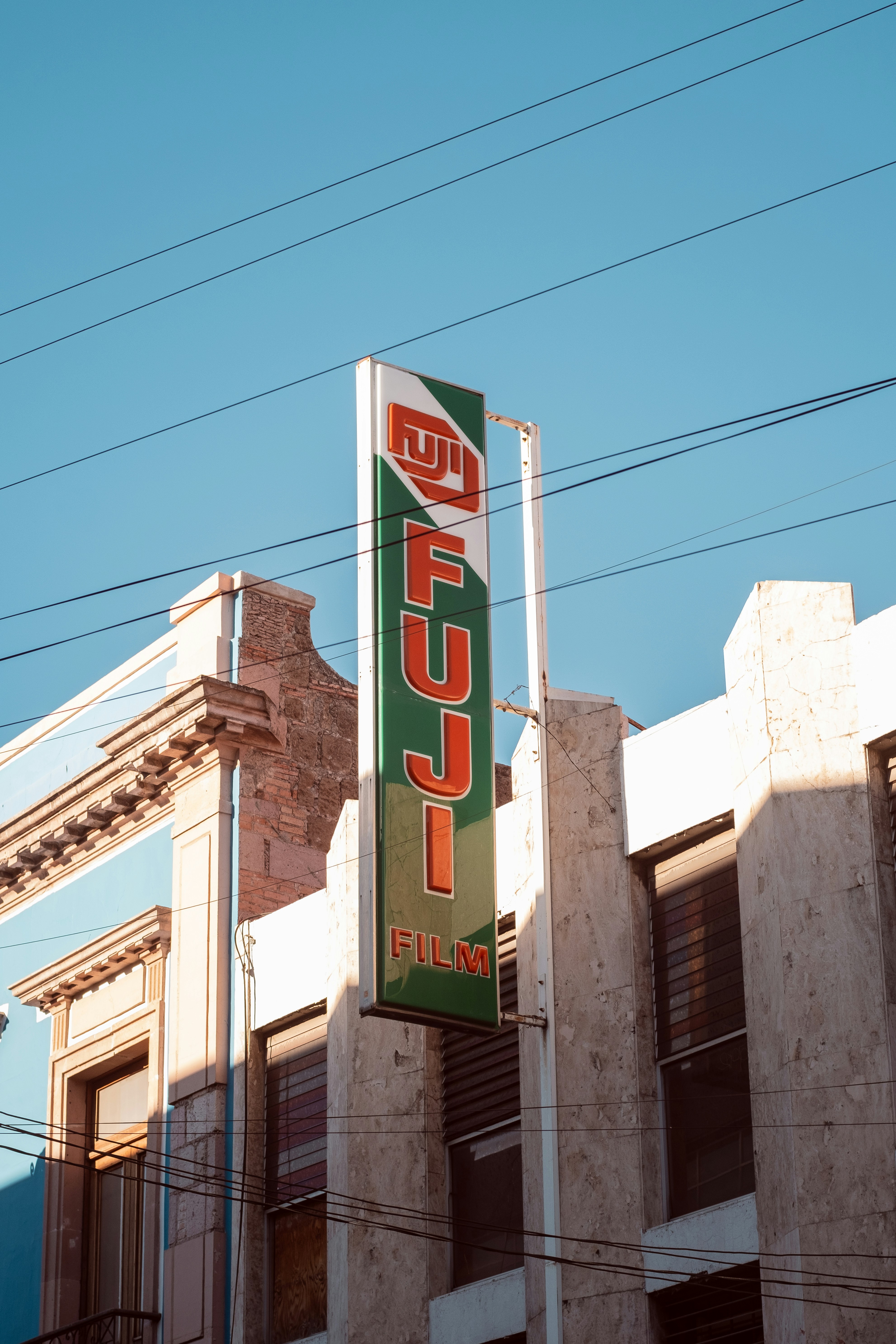 Vertical Fuji Film logo sign rises beside a weathered brick façade under a clear blue sky