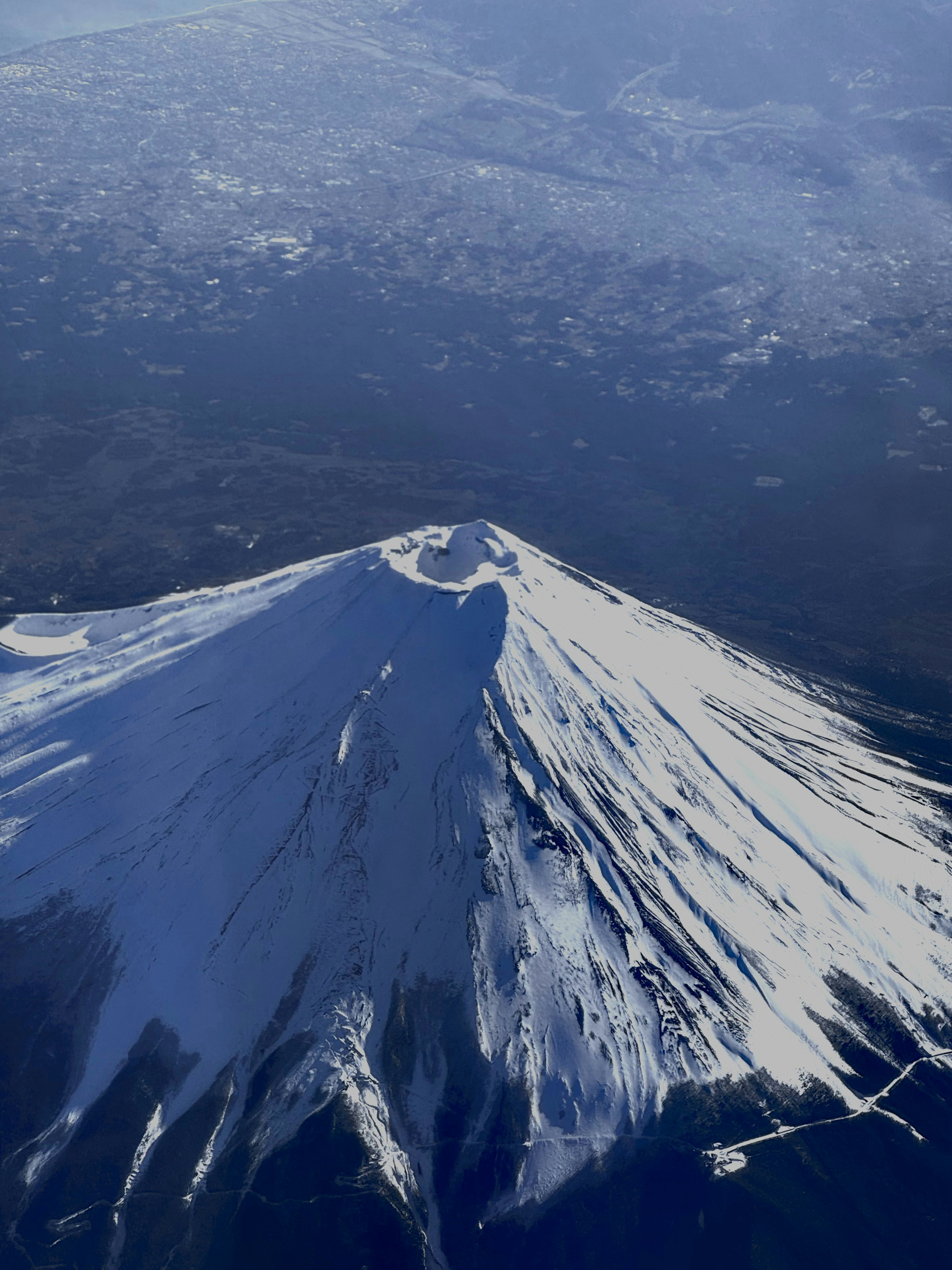 Aerial photograph of a snow-covered volcanic peak viewed from above. Ridges trace down the cone, revealing a stark, glacier-lit summit.