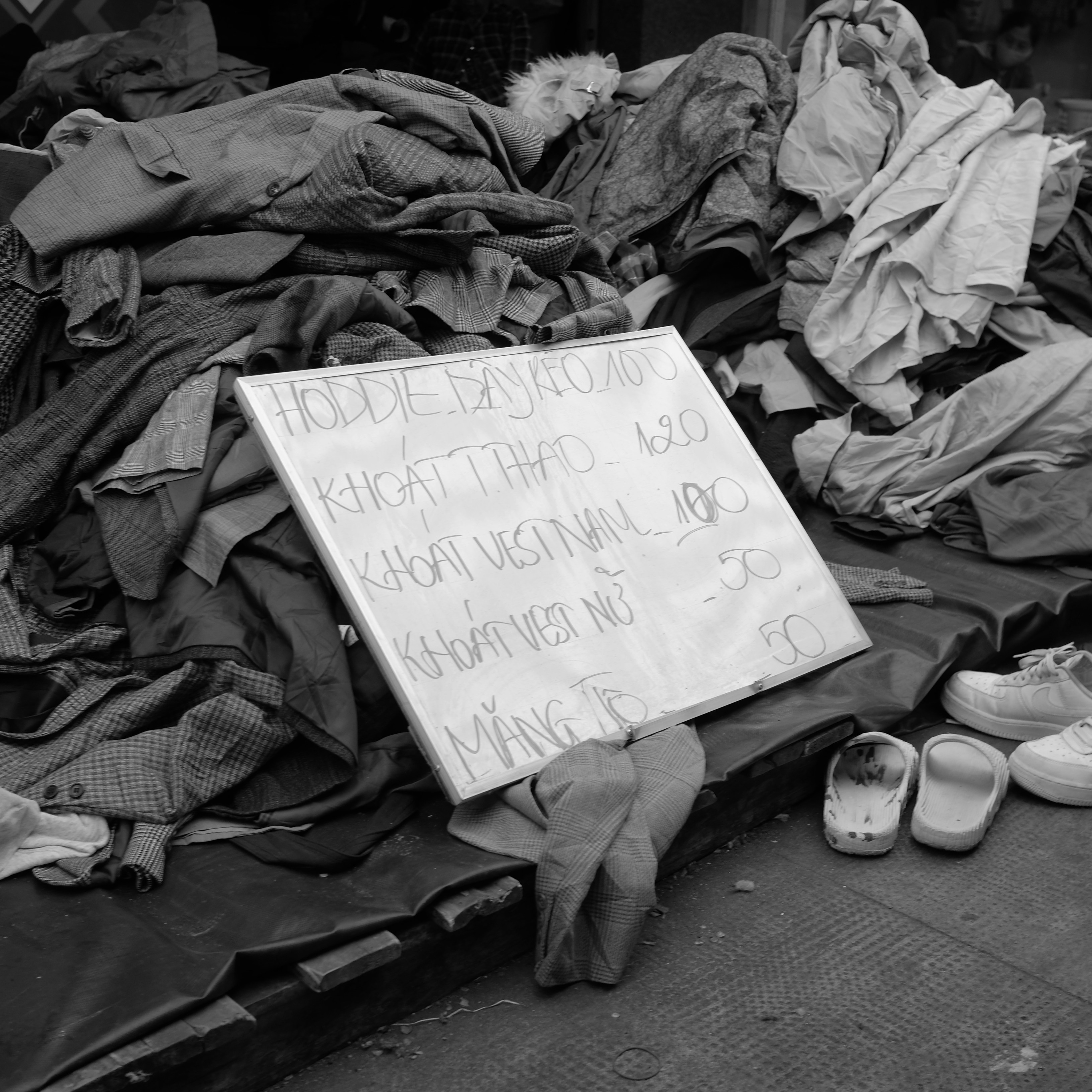 A jumble of discarded clothing and a price list on a sign, hinting at the stories behind each item. The scene captures the essence of a bustling market atmosphere.