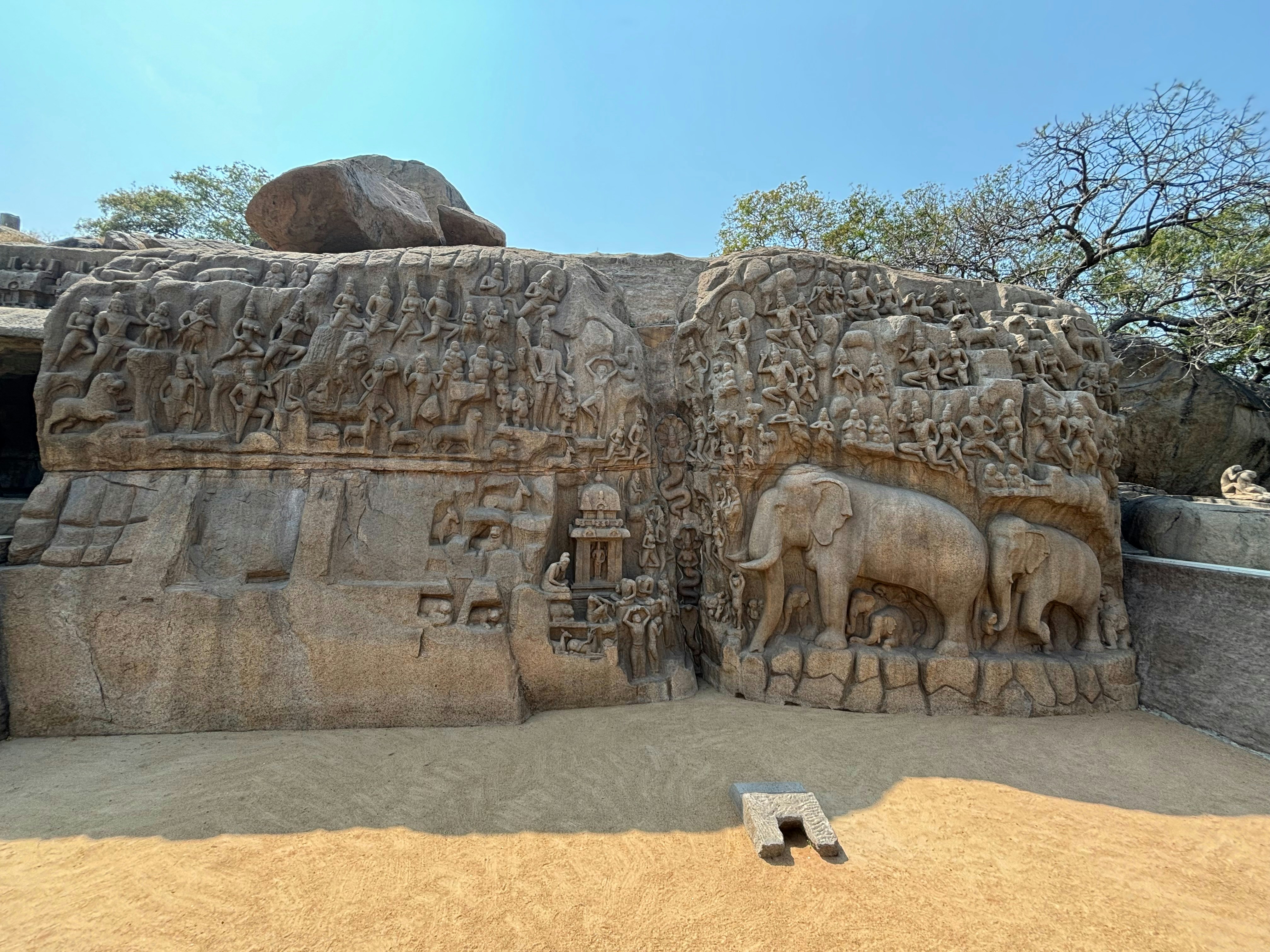 Intricate rock carvings depicting elephants and a myriad of figures, showcasing the artistry of ancient artisans. The scene is set against a backdrop of natural stone and sparse trees.