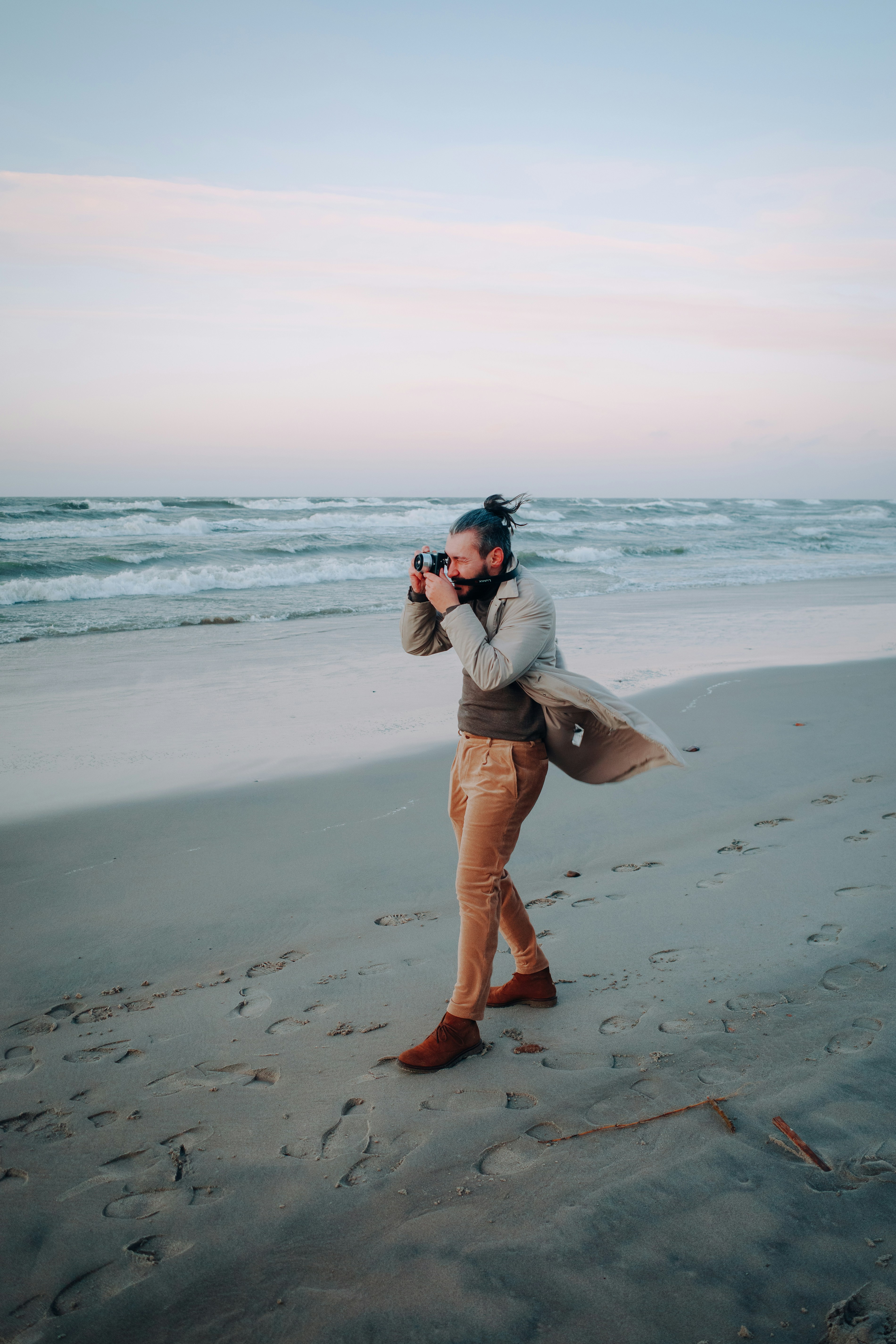 A solitary photographer on a windswept beach raises a camera to the eye as waves roll toward the shore. Footprints mark the damp sand, conveying movement and solitude.