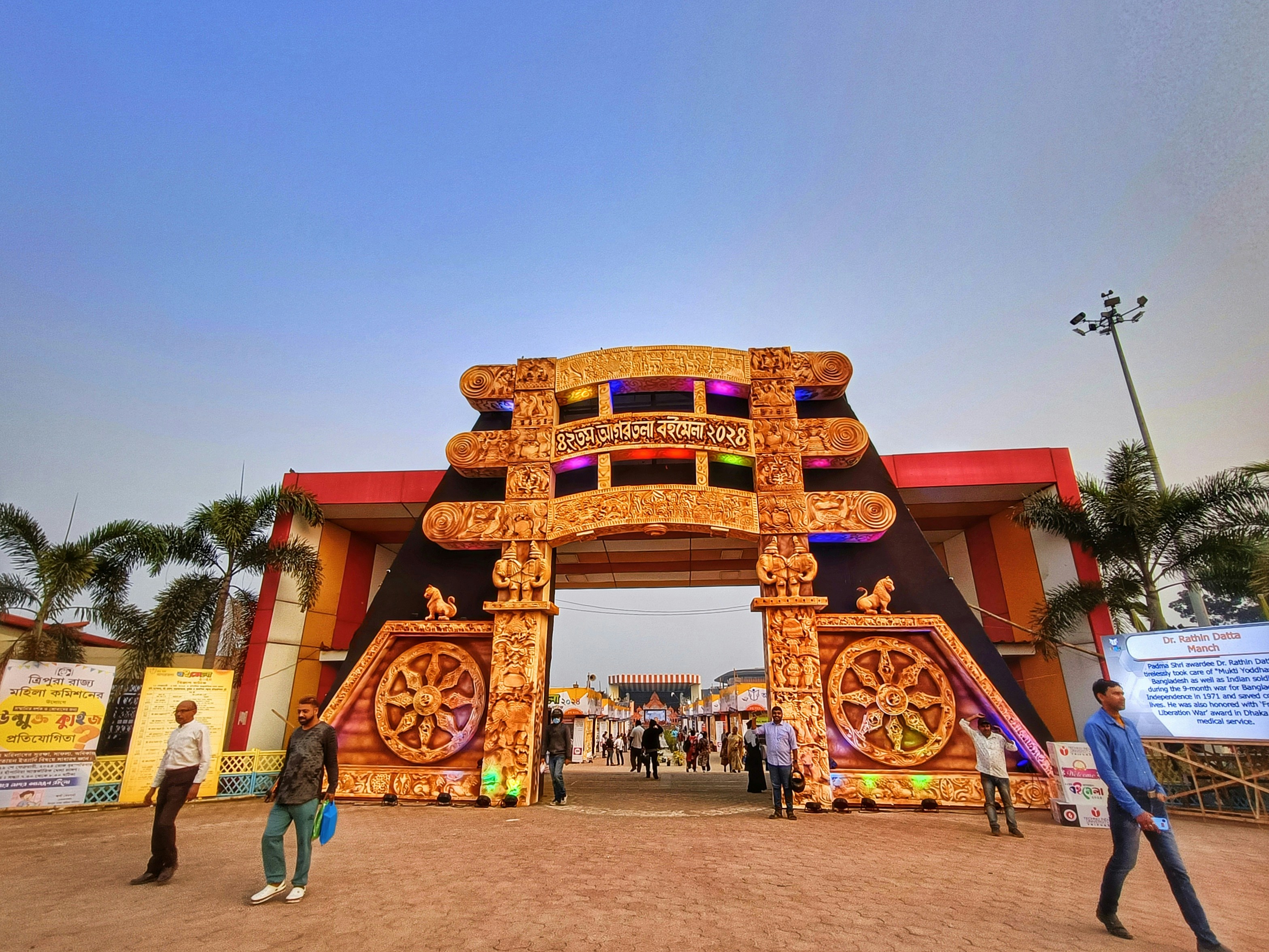 Photograph of an ornate orange gateway arch at a festival entrance with people walking beneath. The focus is the grand arch and the welcoming passage.