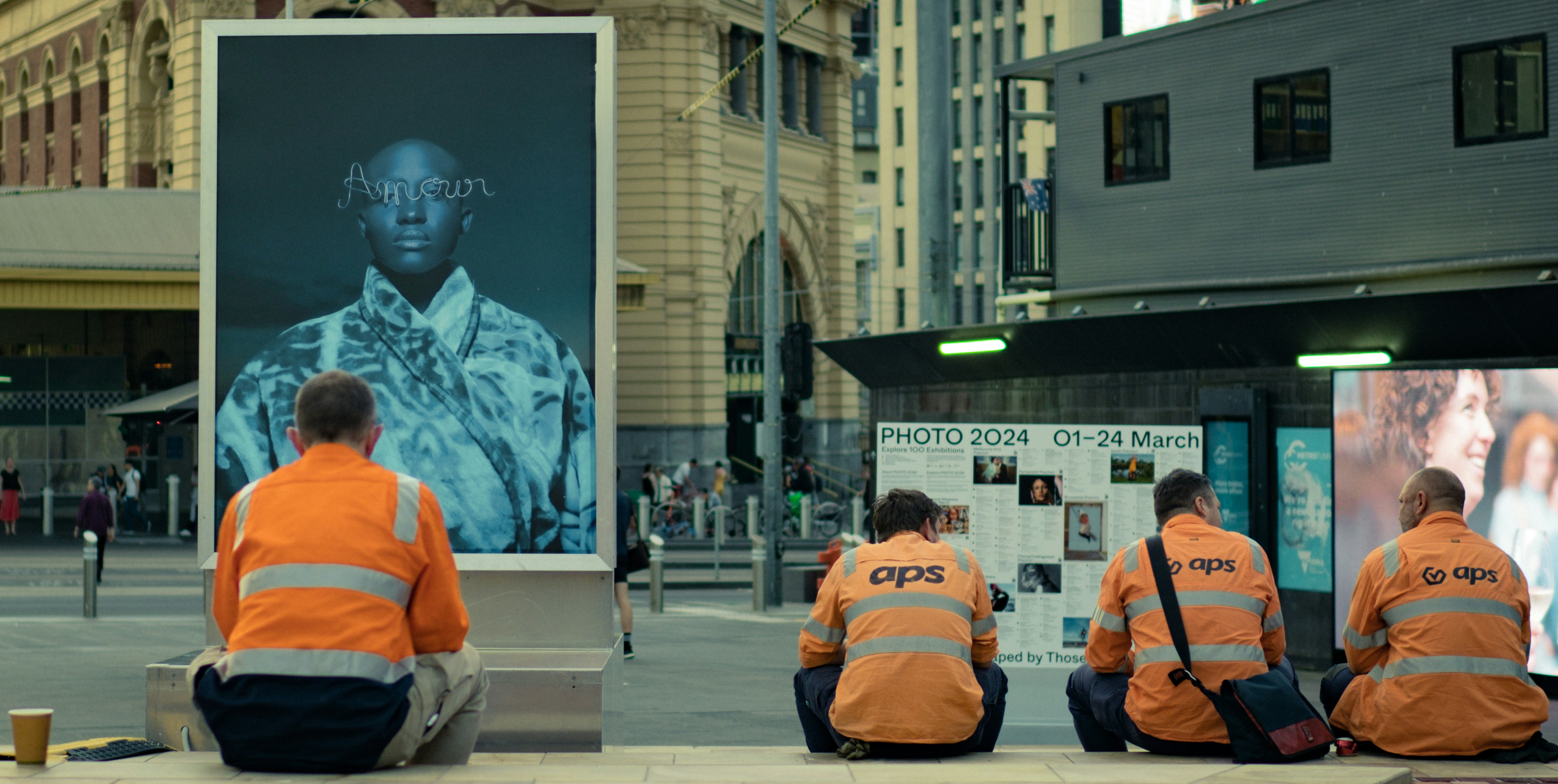 Workers in orange vests seated near a public art display on a city street.