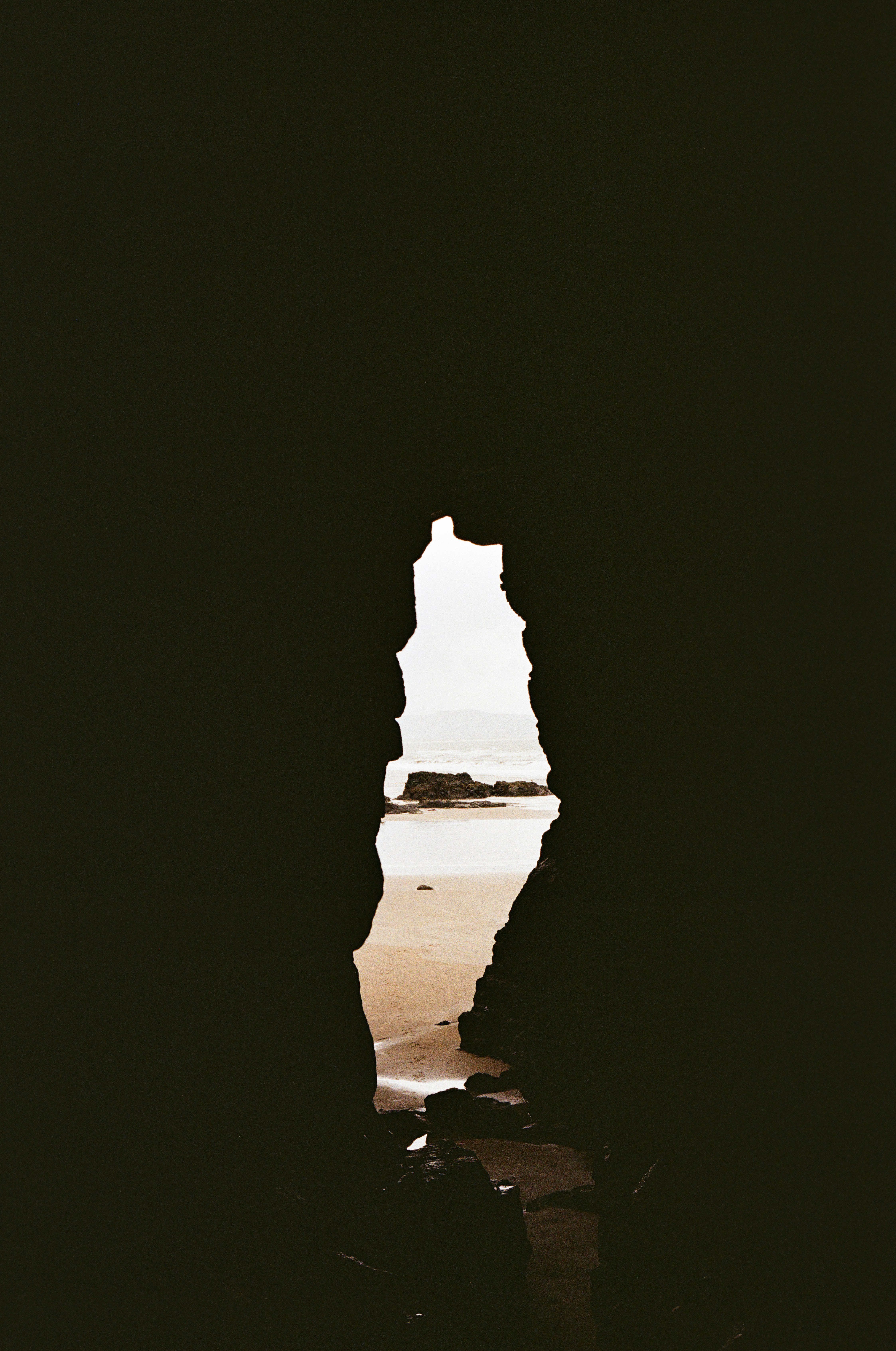 A jagged cave opening frames a sunlit beach and sea beyond, captured in a stark, high-contrast photograph.