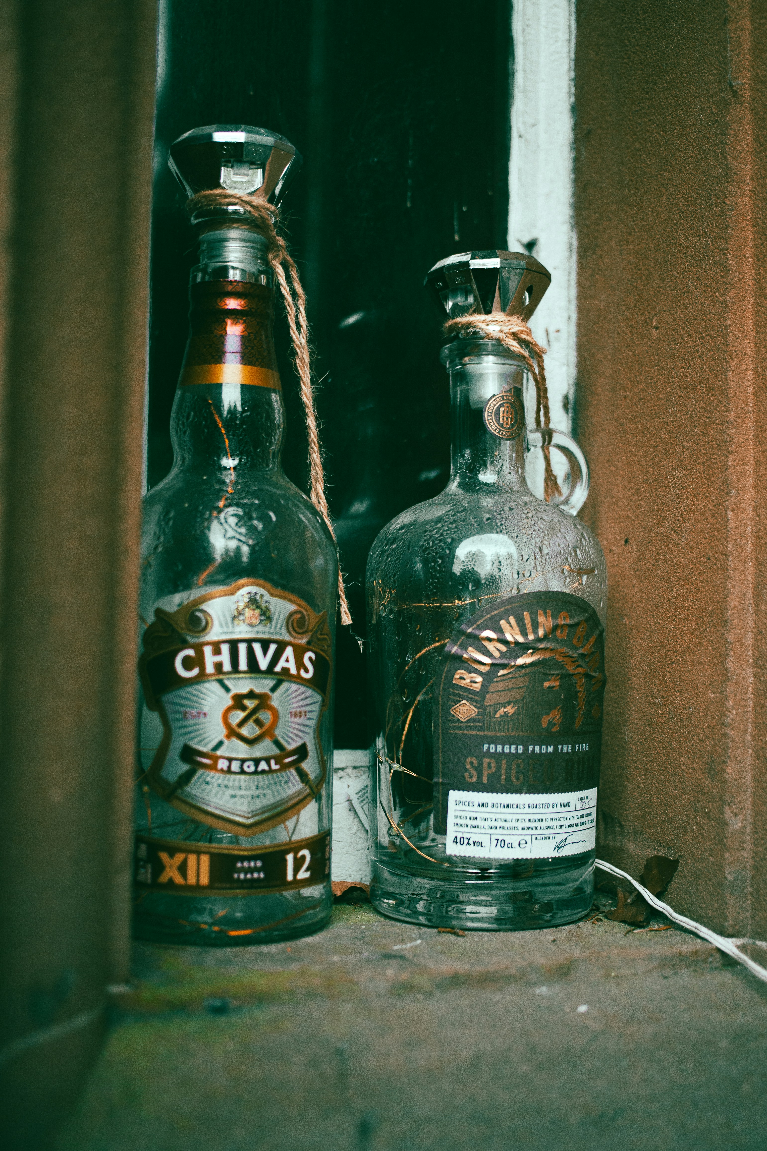 Two glass liquor bottles, Chivas Regal and a spiced-label bottle, rest side-by-side in a narrow, dusty cabinet. The moody composition emphasizes weathered glass, rope closures, and detailed labels.